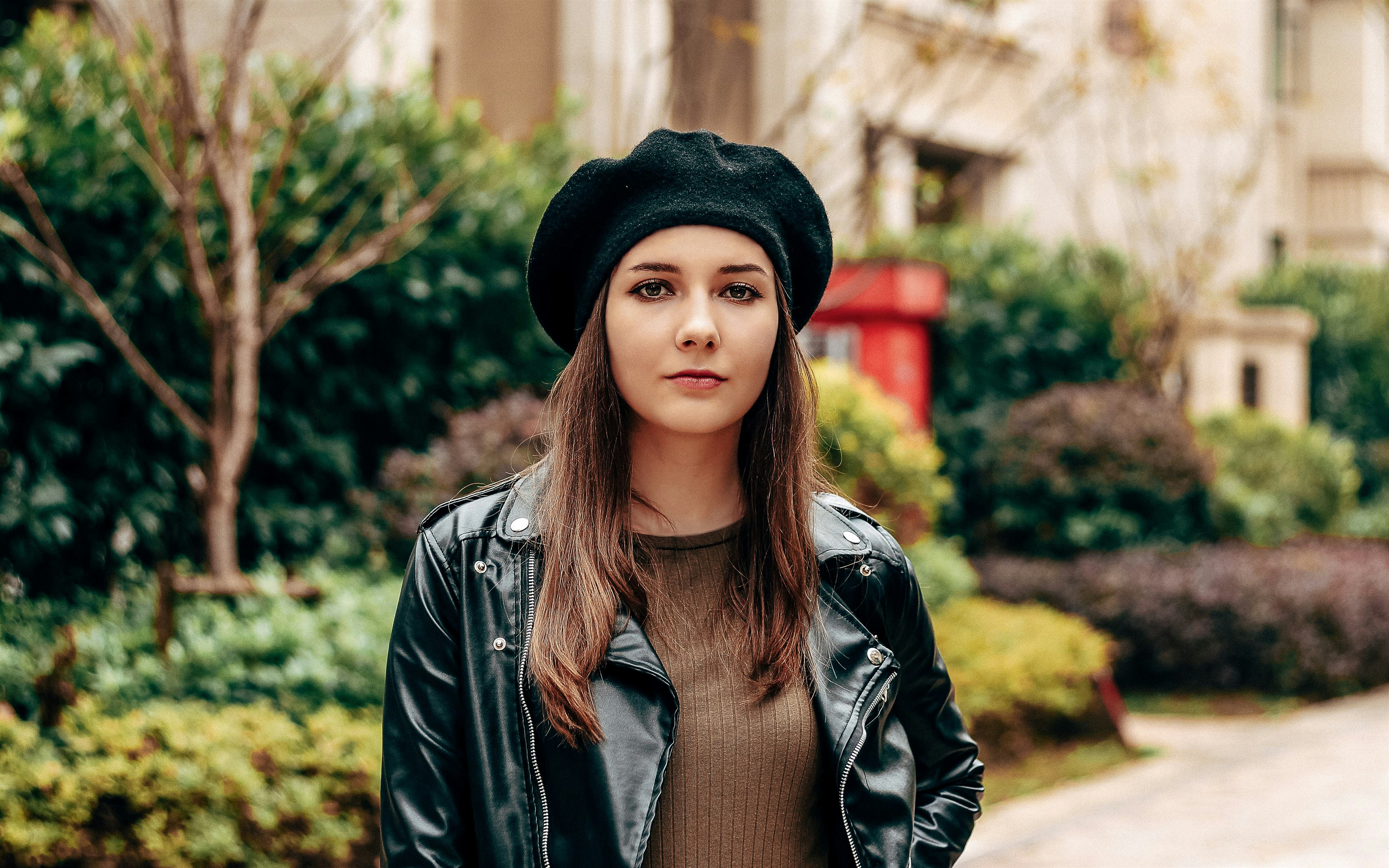 A young woman in a black beret and leather jacket stands confidently amidst lush greenery in an urban setting.