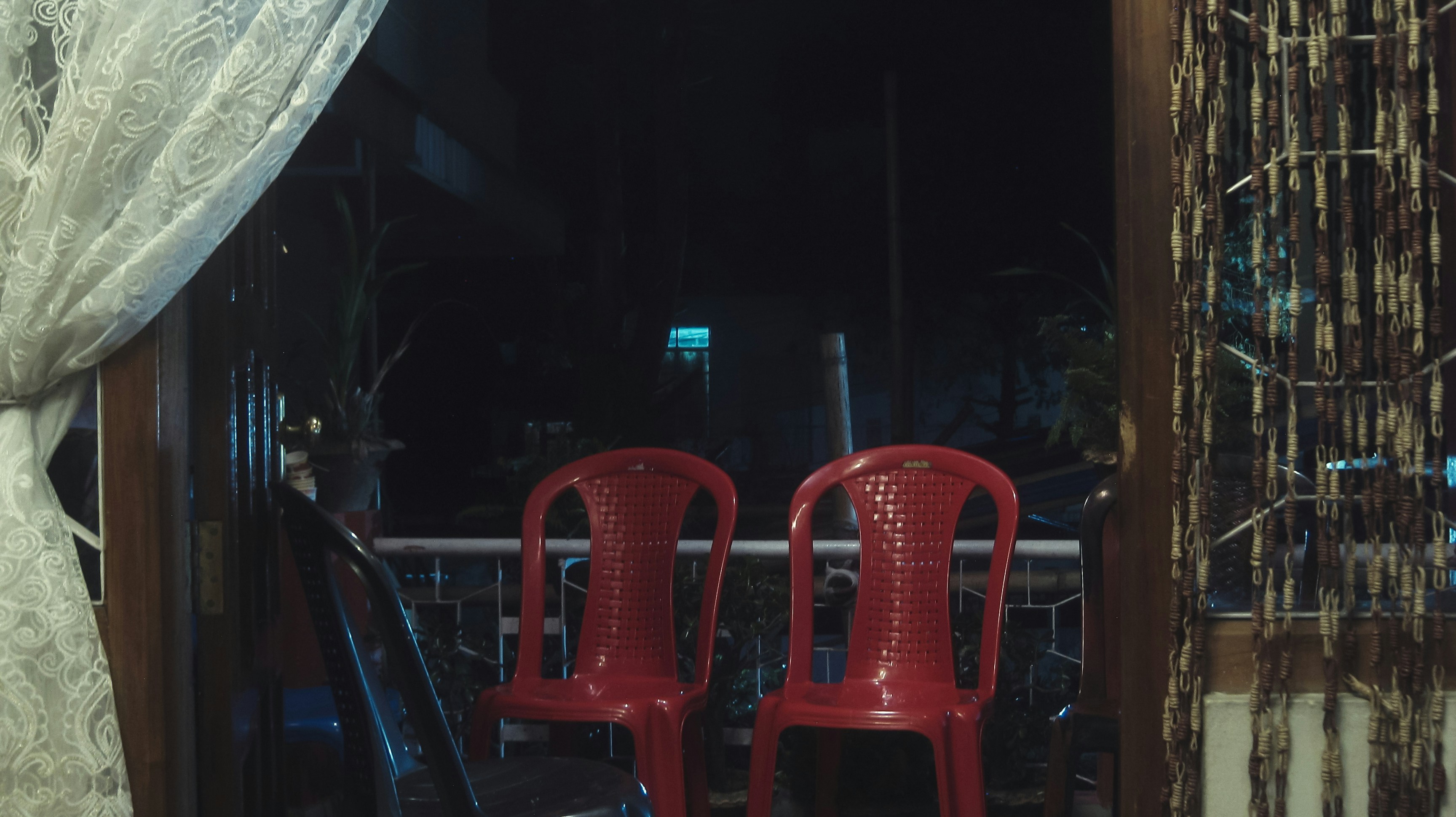 Two red plastic chairs sit on a dim porch beneath lace curtains. A dark doorway and faint blue light recede into the background.