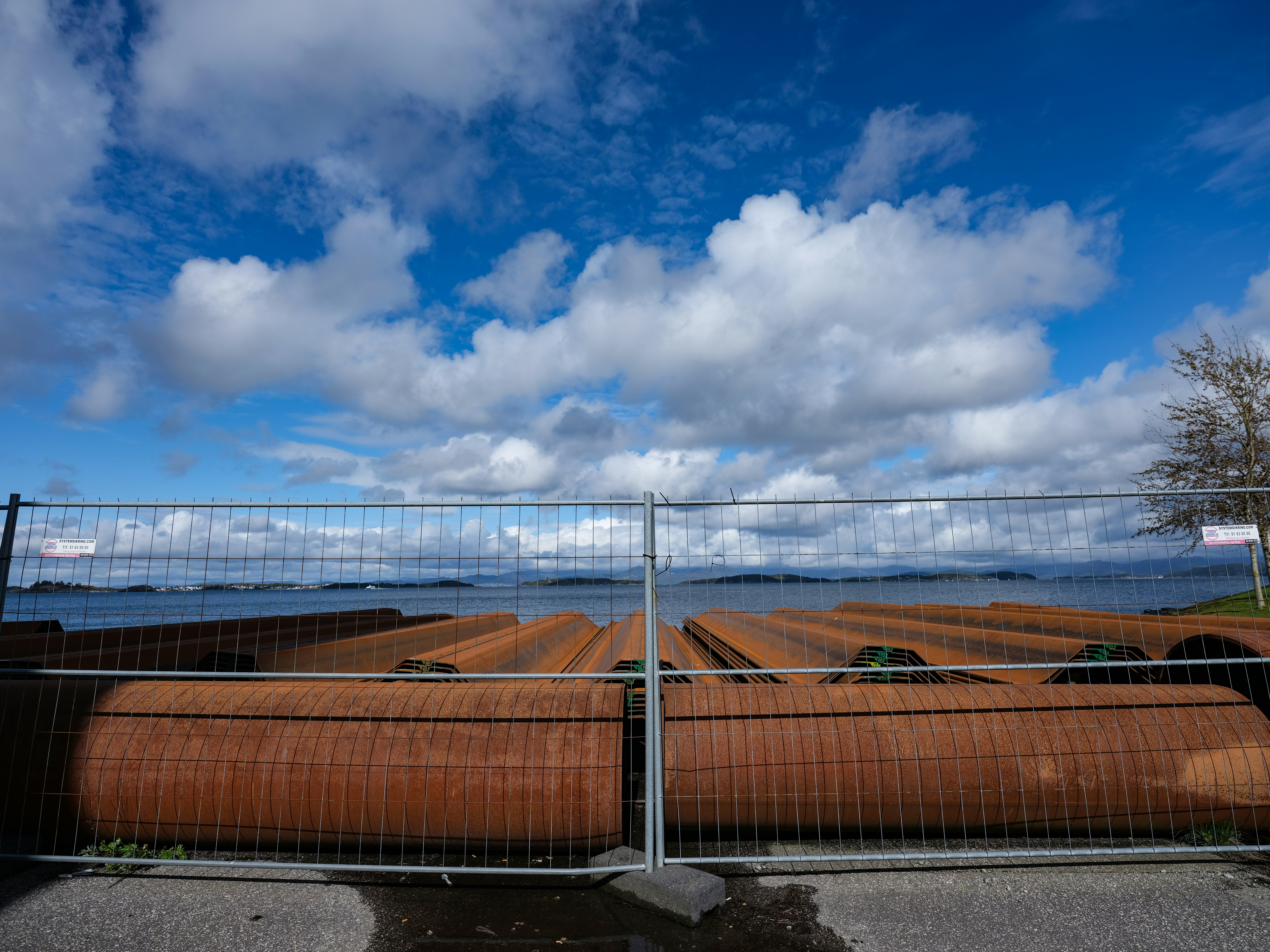 Agricultural fields bordered by a metal fence under a partly cloudy sky, with distant ocean views.