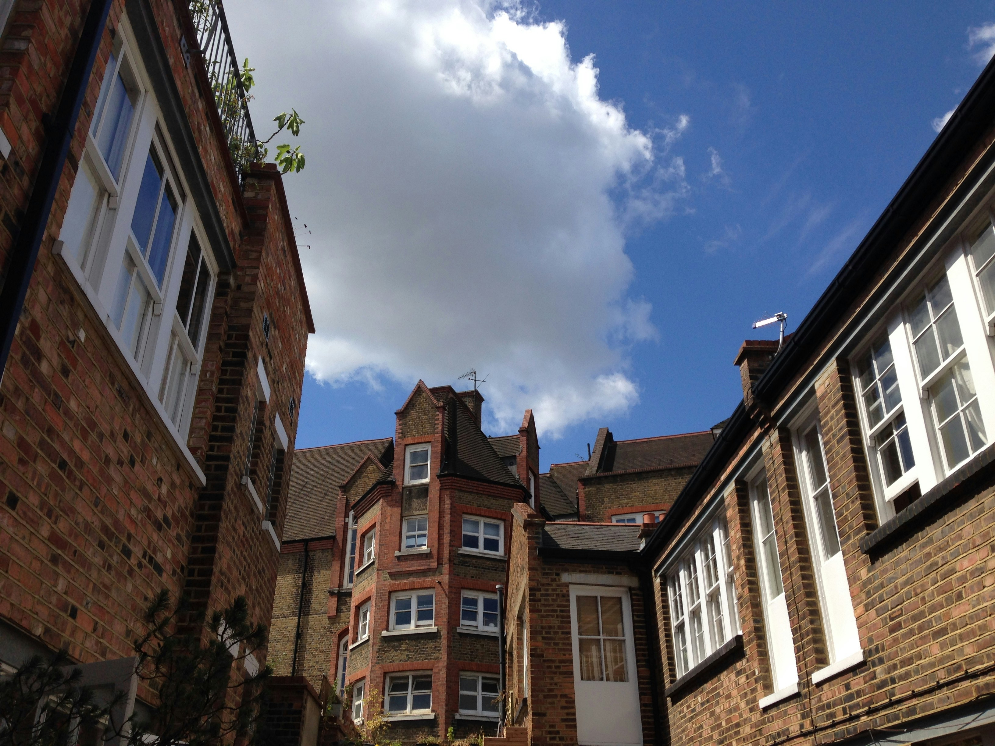 Brick buildings flank a narrow courtyard beneath a bright blue sky, with a rounded central tower anchoring the scene. The composition emphasizes brick textures and architectural rhythm.
