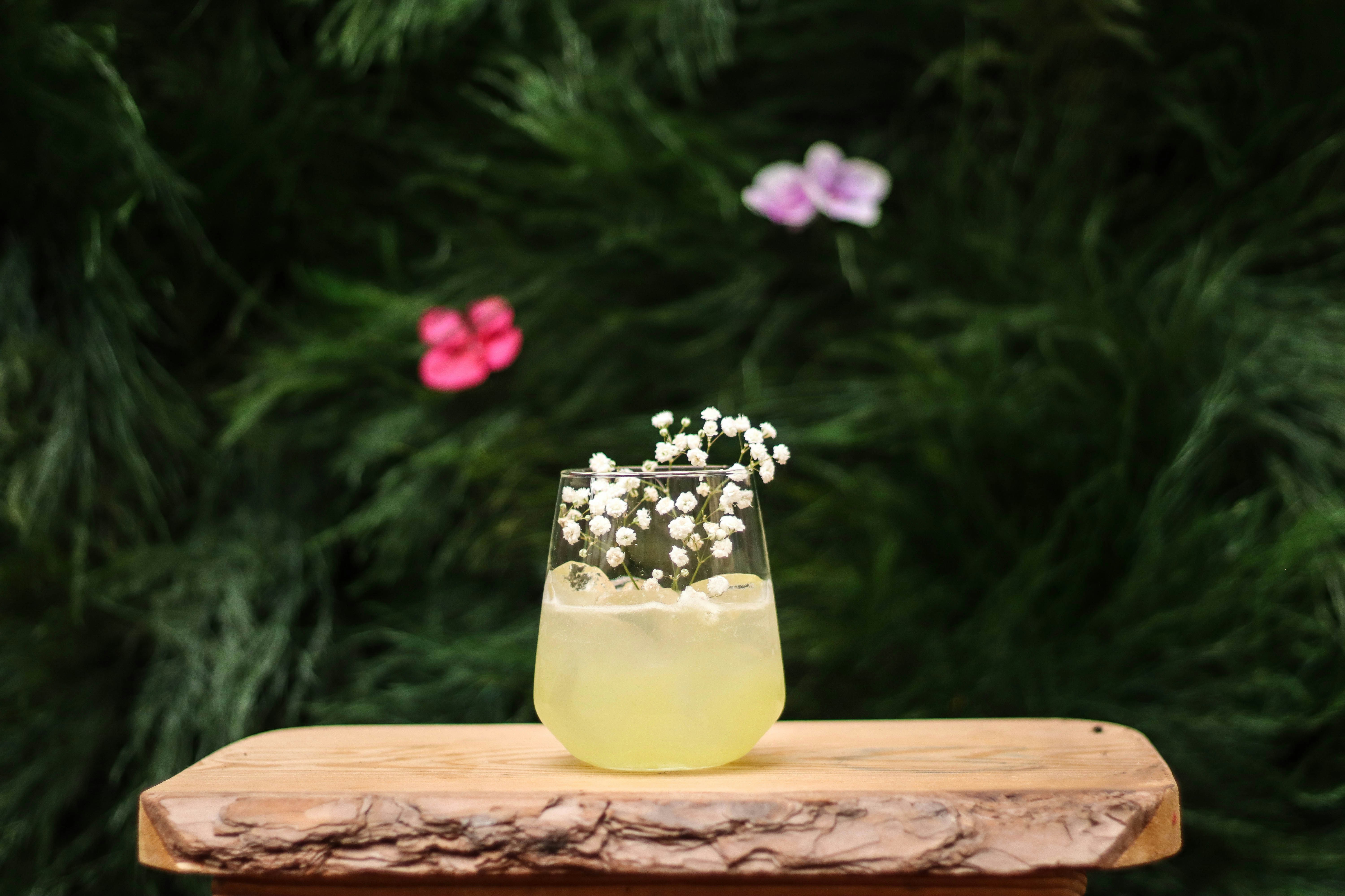 Delicate white flowers garnish a pale yellow drink on a wooden surface, set against a lush green backdrop with soft pink and red blooms.