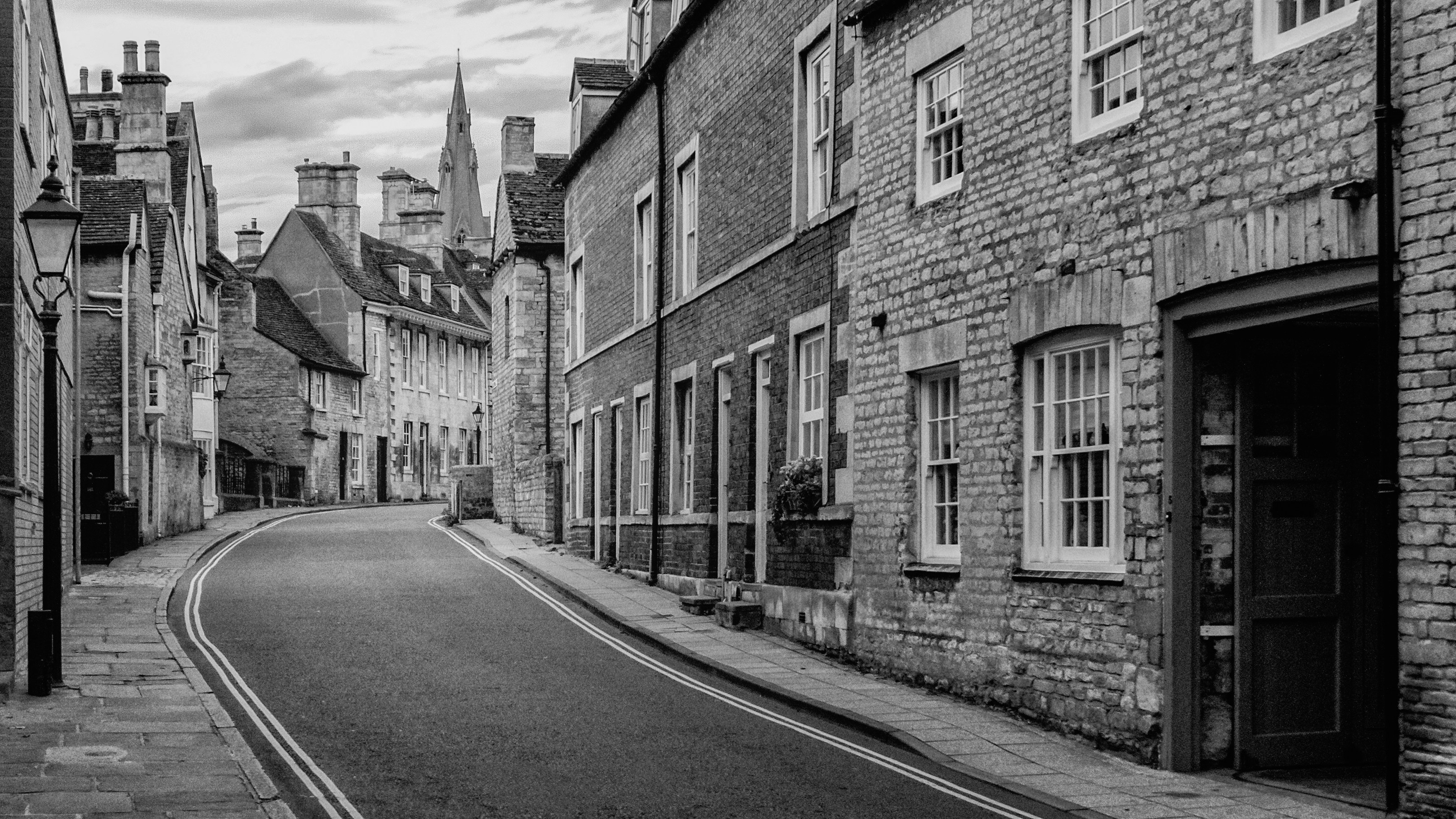 Charming narrow street lined with historic stone buildings, leading towards a distant steeple under a cloudy sky.