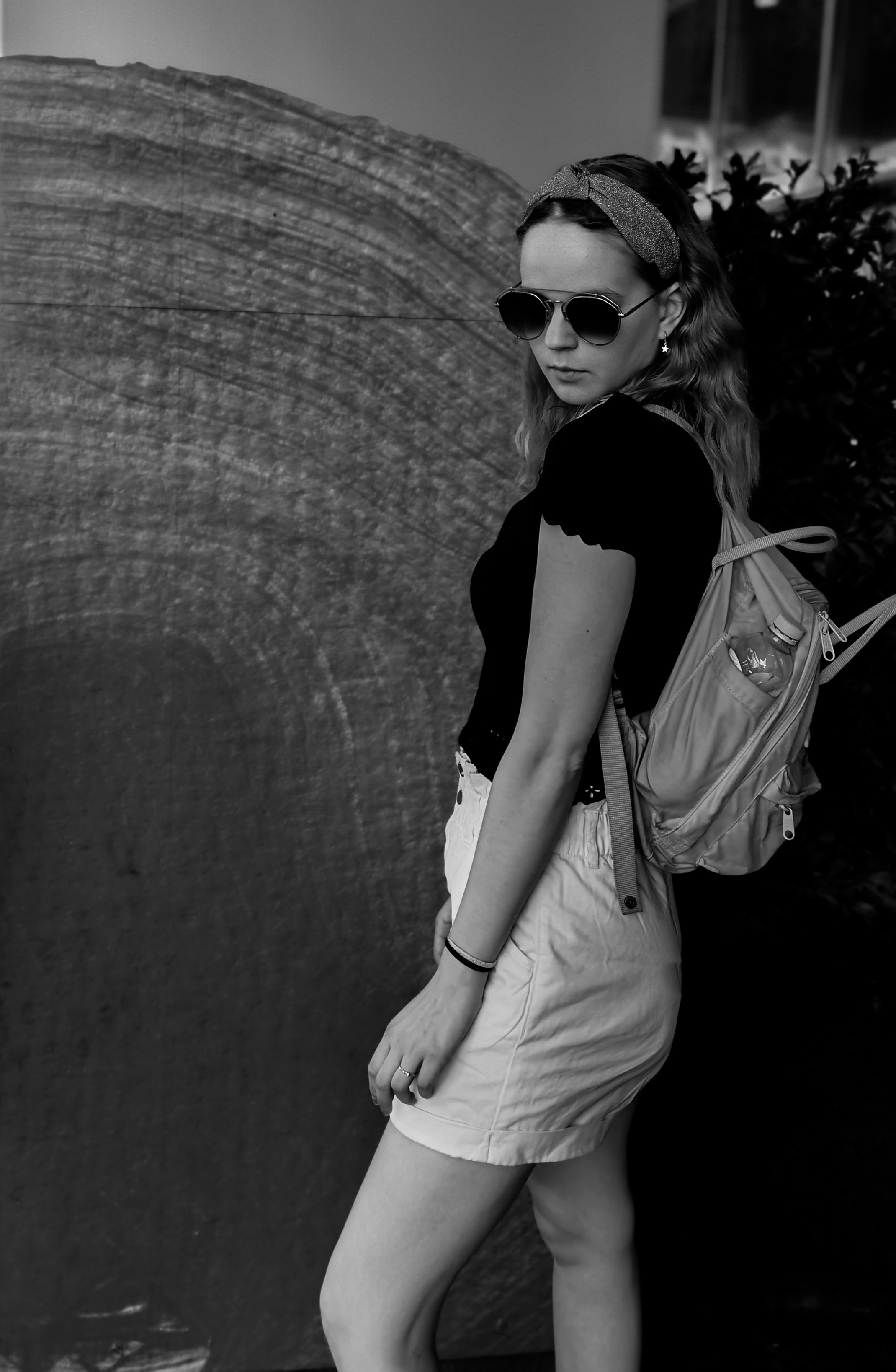Black-and-white street portrait of a woman wearing sunglasses and a backpack, posed beside a large curved textured surface. The composition emphasizes contrast and a poised, urban mood.