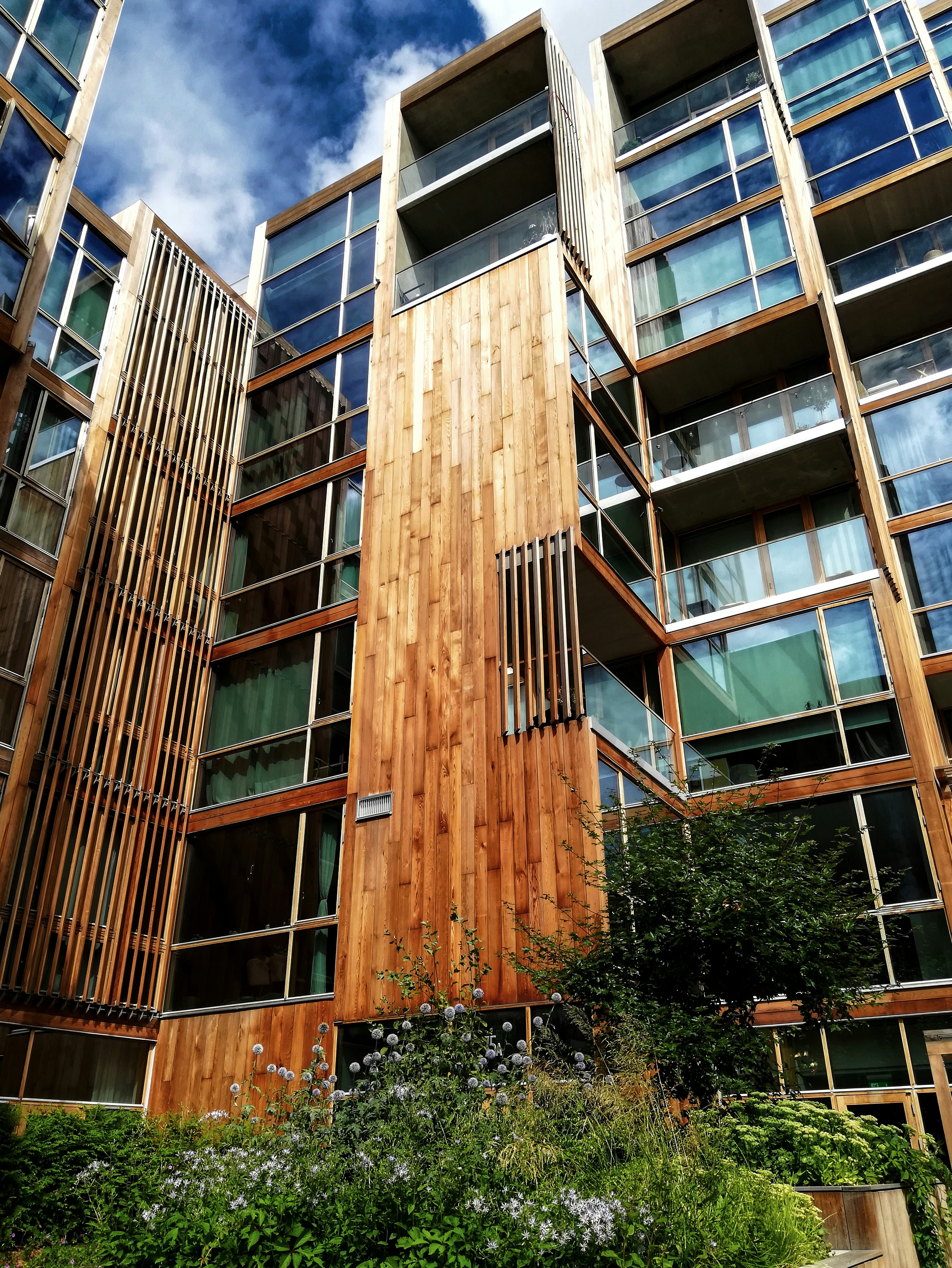 Modern wooden facade of an apartment building surrounded by lush greenery and a blue sky with scattered clouds.