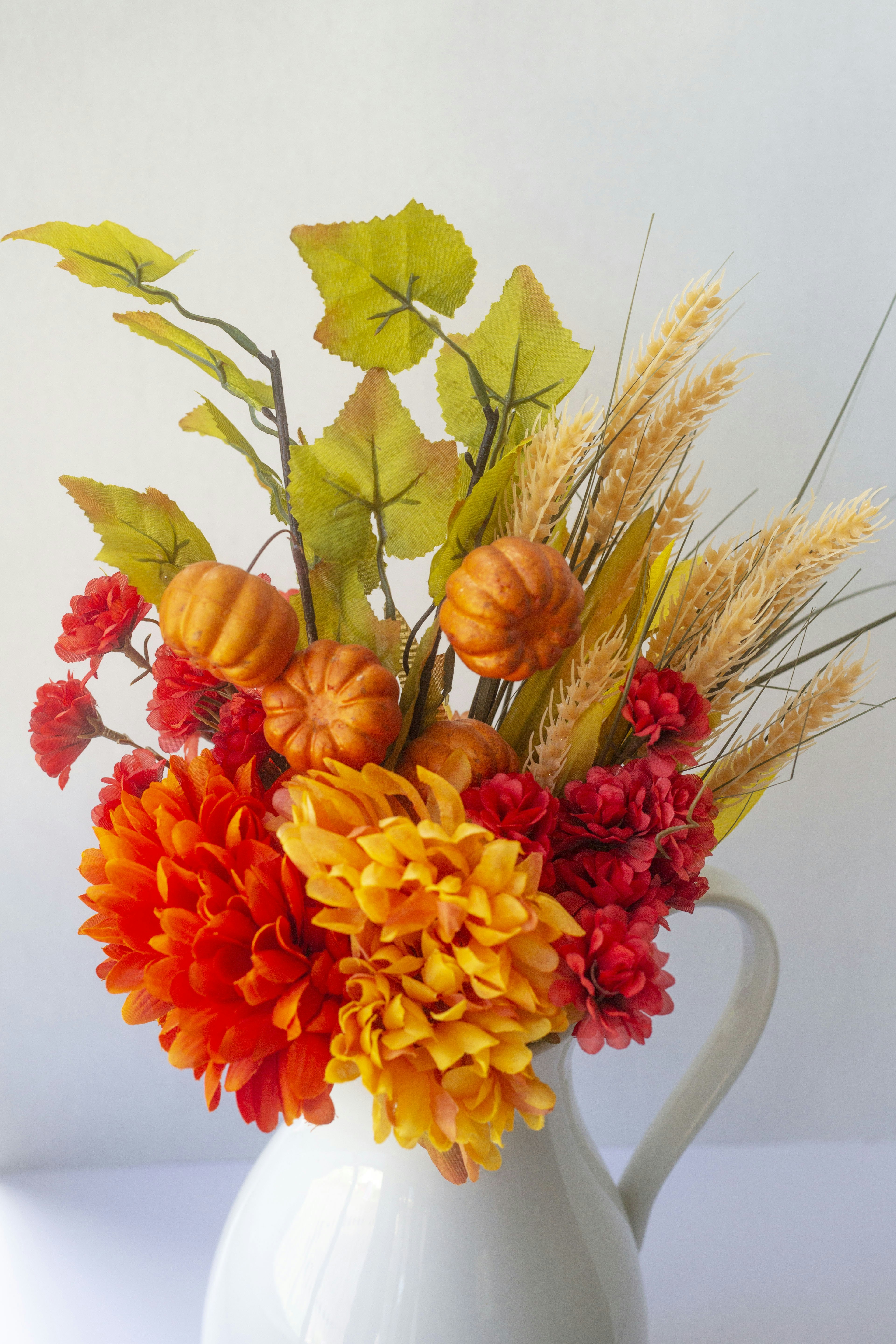 Vibrant bouquet featuring artificial flowers, pumpkins, and wheat, arranged in a white pitcher. The composition celebrates the colors and textures of fall.