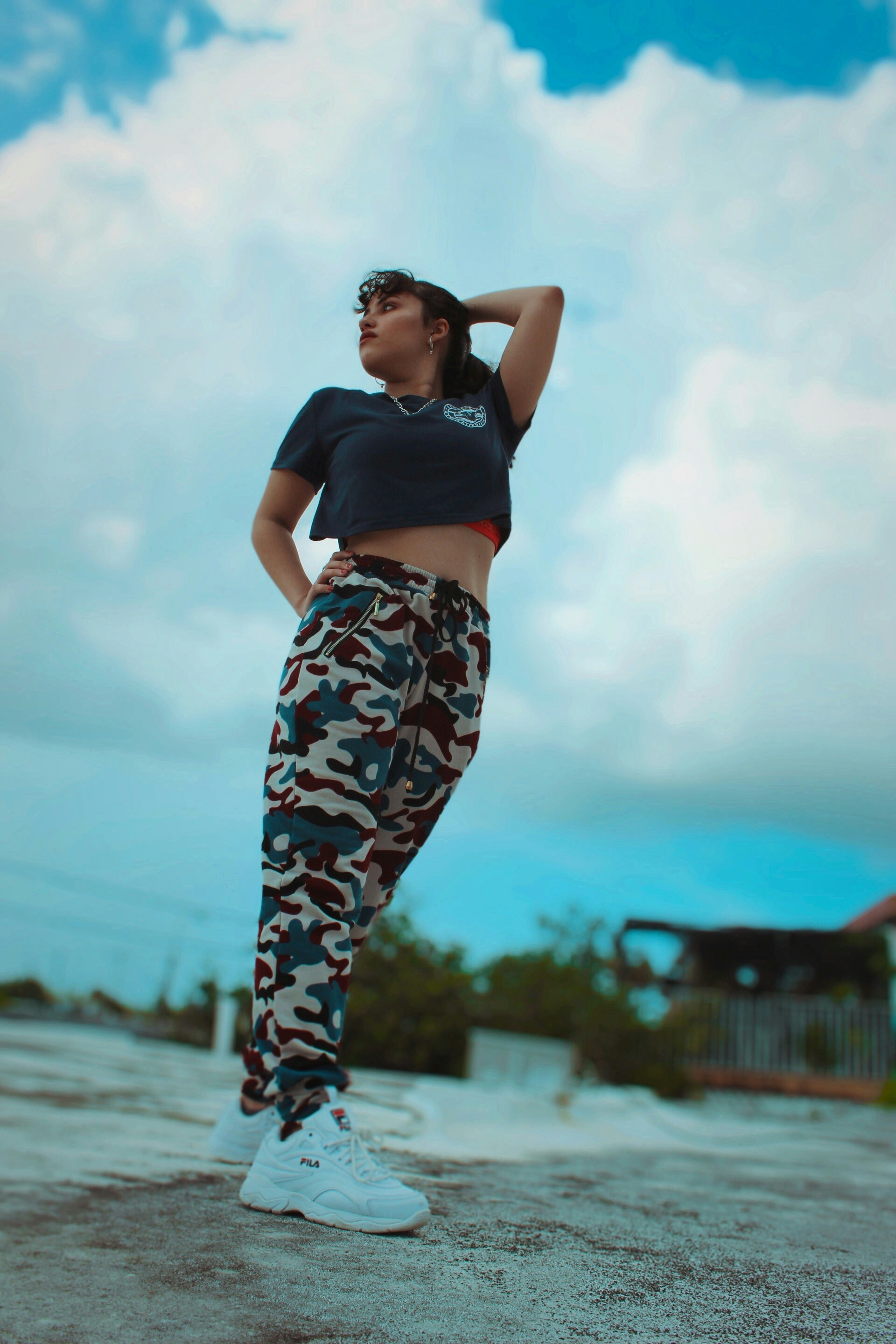 Young woman posing confidently in camouflage pants and a cropped top against a backdrop of dramatic clouds.
