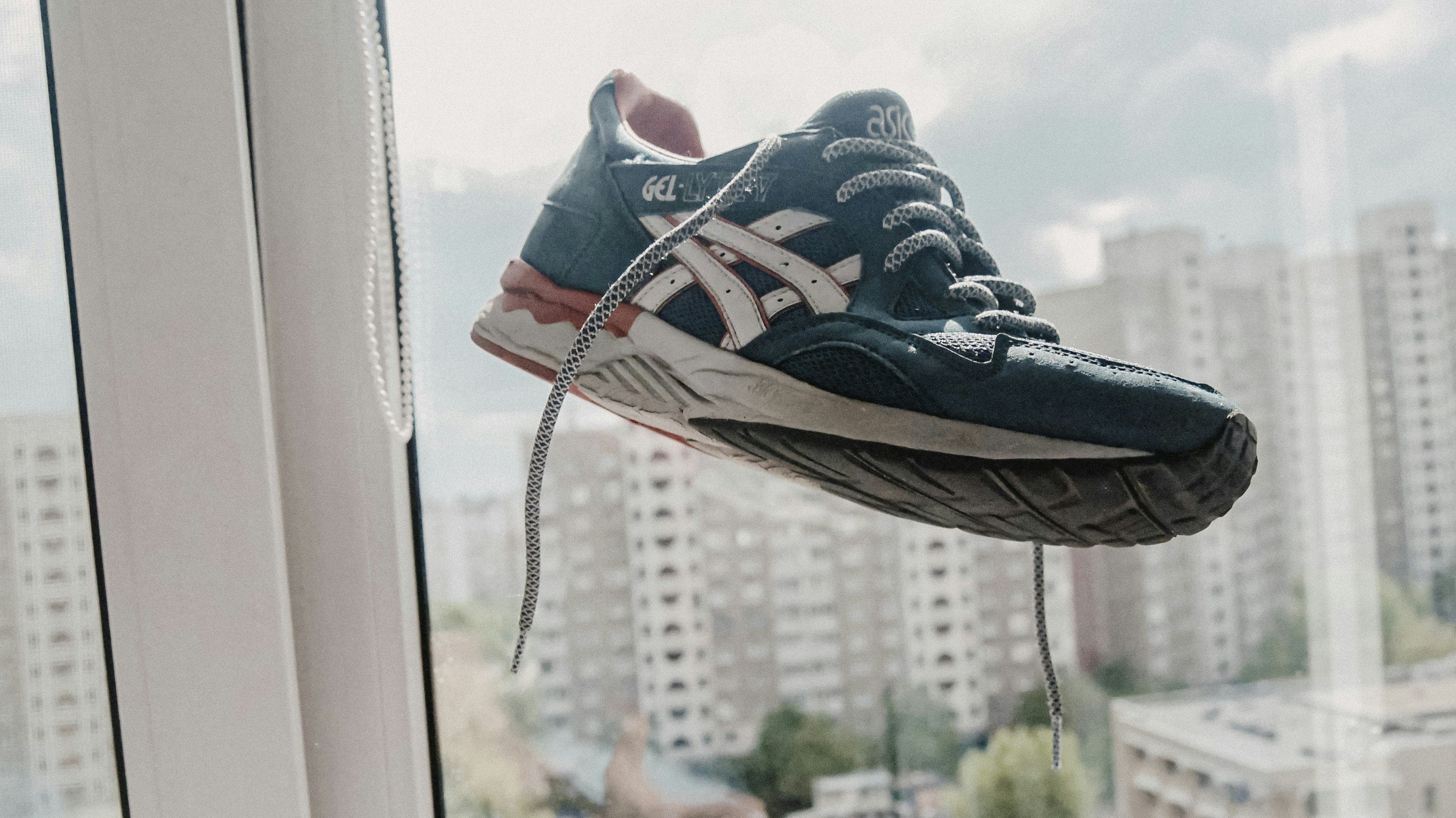A single sneaker suspended in mid-air near a window, with a cityscape backdrop. The scene captures a moment of urban lifestyle.