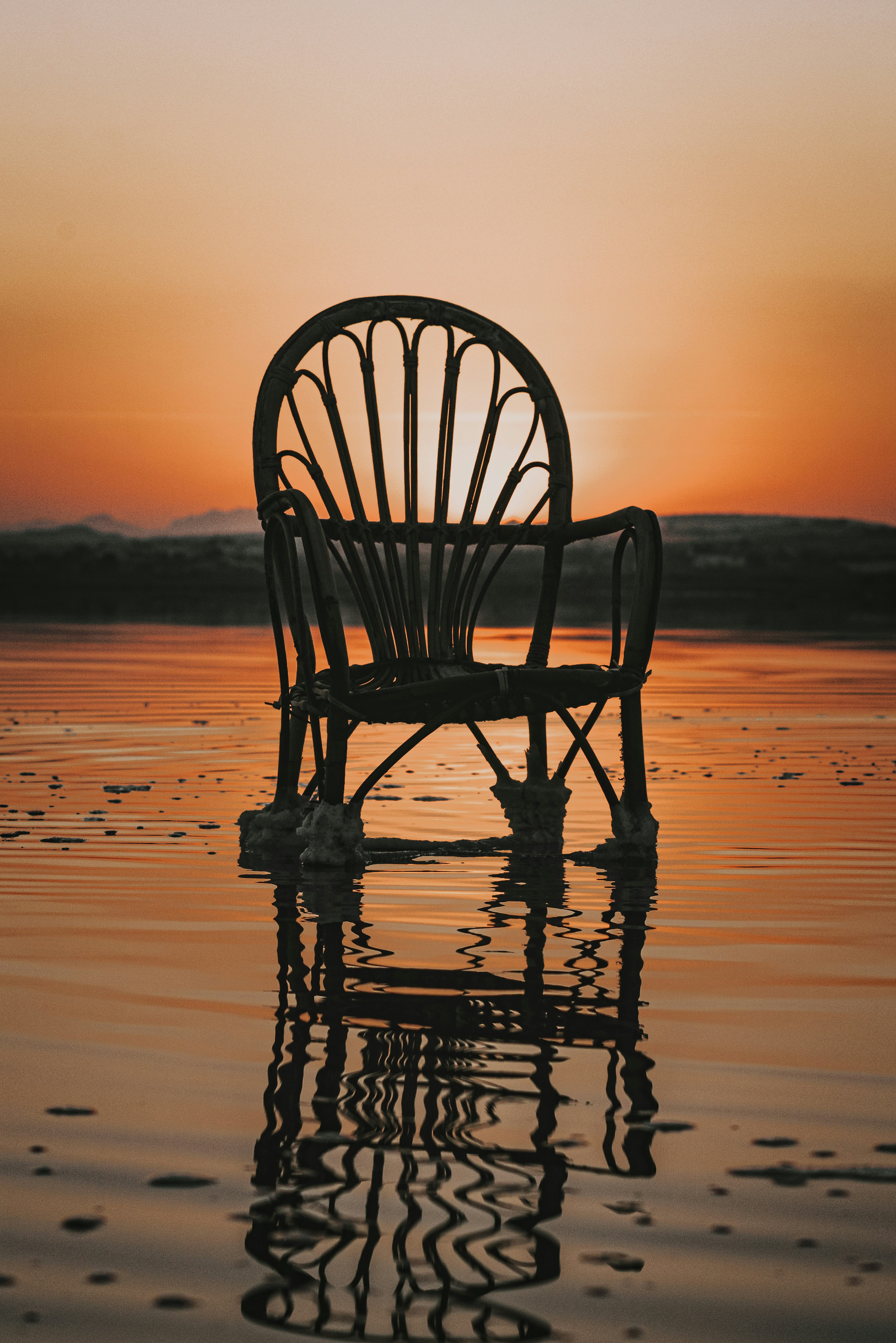 A solitary chair stands in shallow water during sunset, creating a striking silhouette against the colorful sky. The calm surface reflects the chair, enhancing the scene's tranquility.