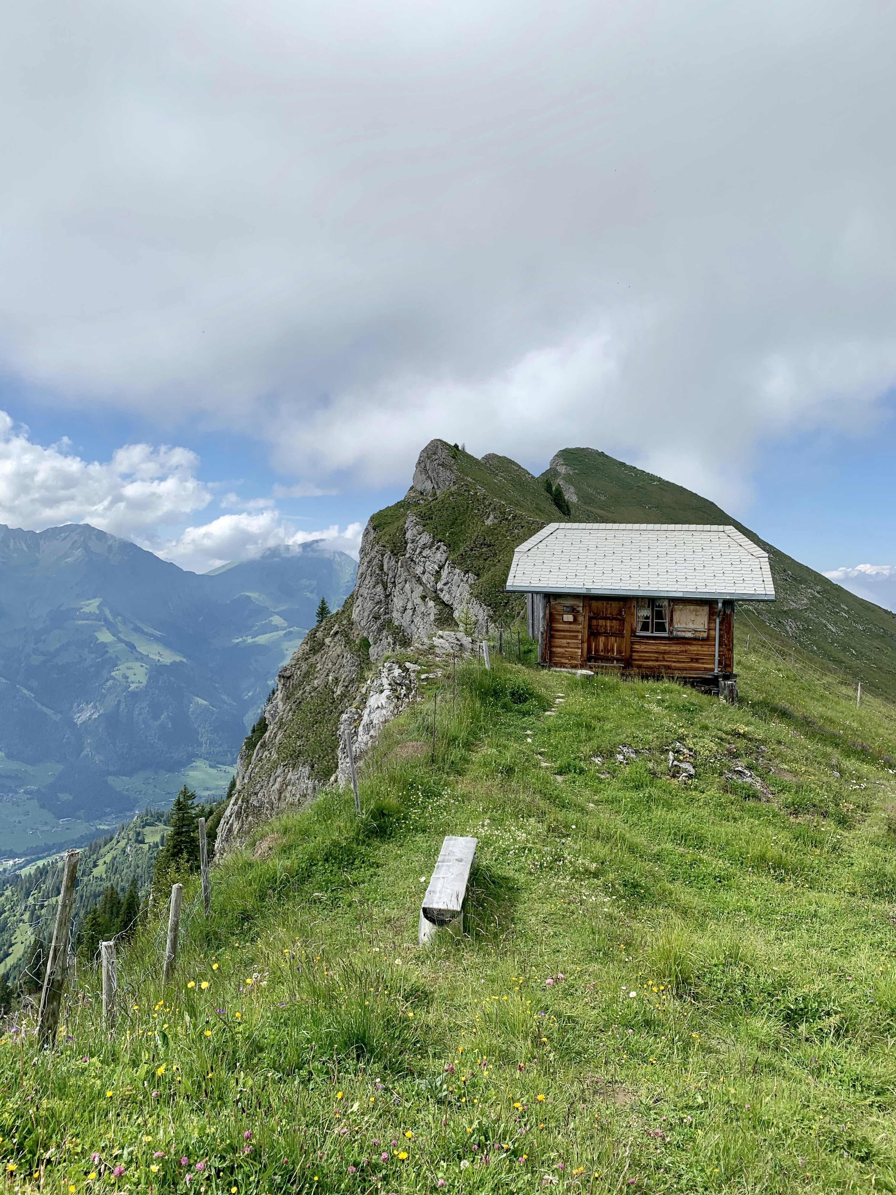 Wooden cabin perched on a mountain ridge, surrounded by lush greenery and distant peaks under a partly cloudy sky.