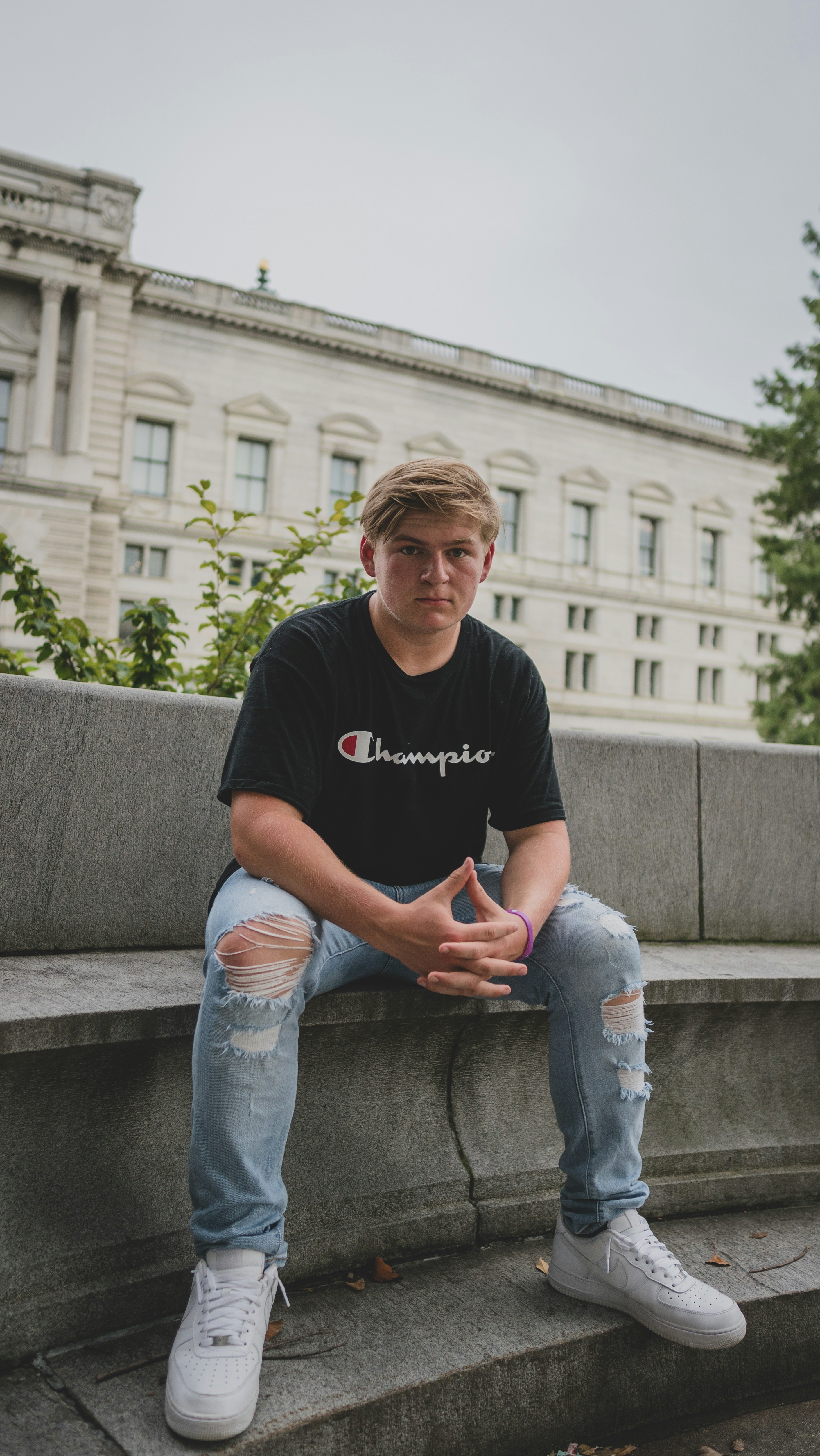 A young man sits on a stone bench, dressed in casual attire, with a historic building in the background, conveying a sense of reflection and urban tranquility.