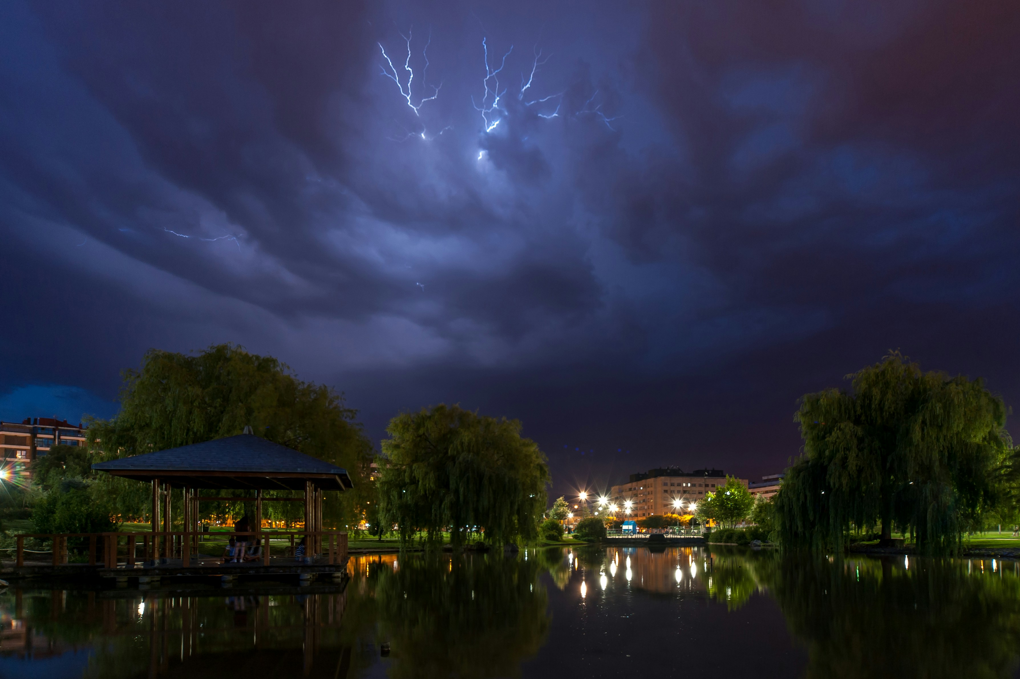 Lightning illuminates a dark sky above a tranquil pond, surrounded by lush greenery and a gazebo, capturing the juxtaposition of nature and urban life.