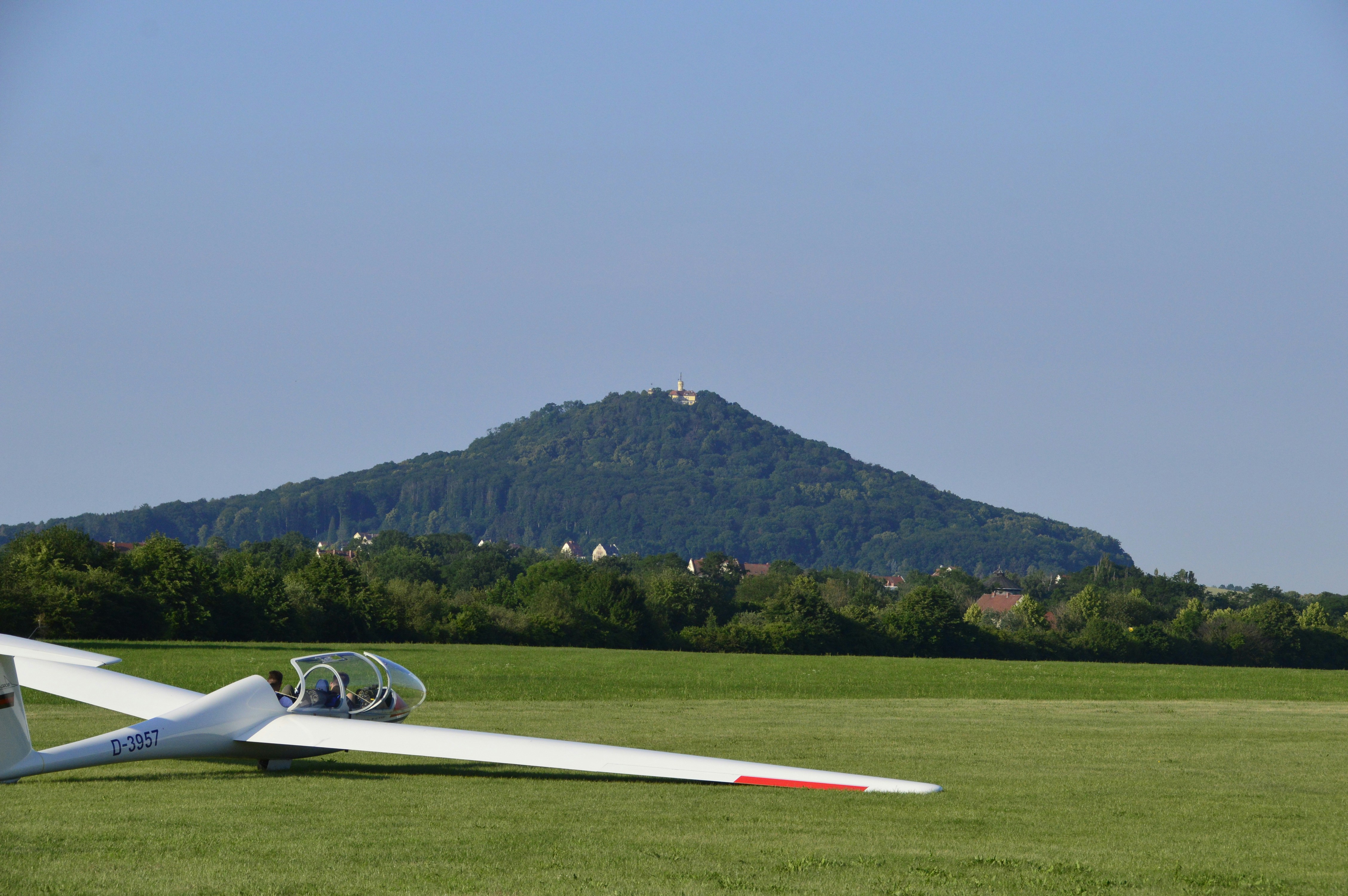 A glider rests on a lush green field, with a distant mountain peak crowned by a small structure under a clear blue sky.
