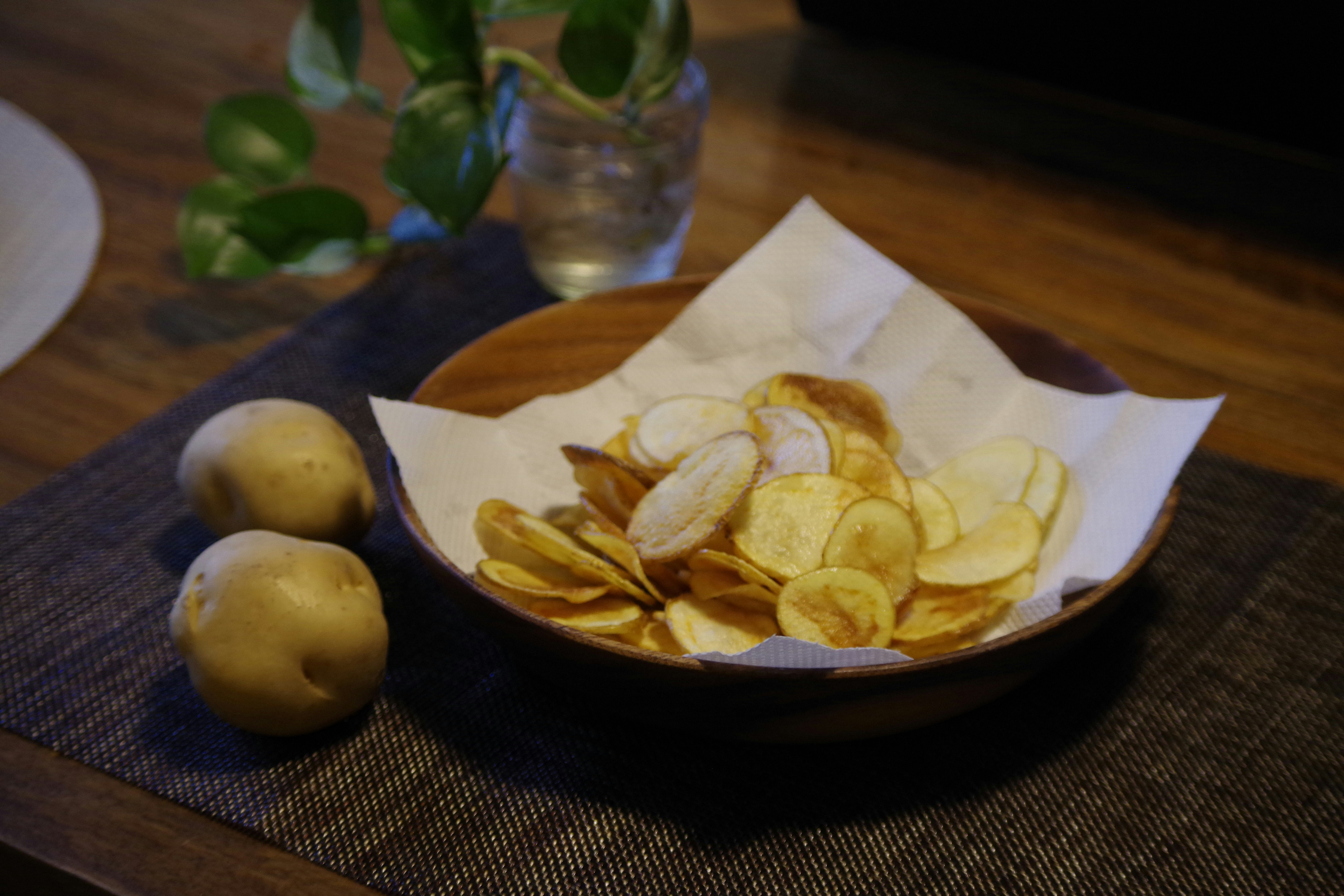A wooden bowl filled with freshly made potato chips rests on a textured mat, accompanied by two whole potatoes and a small glass of water with a green plant.