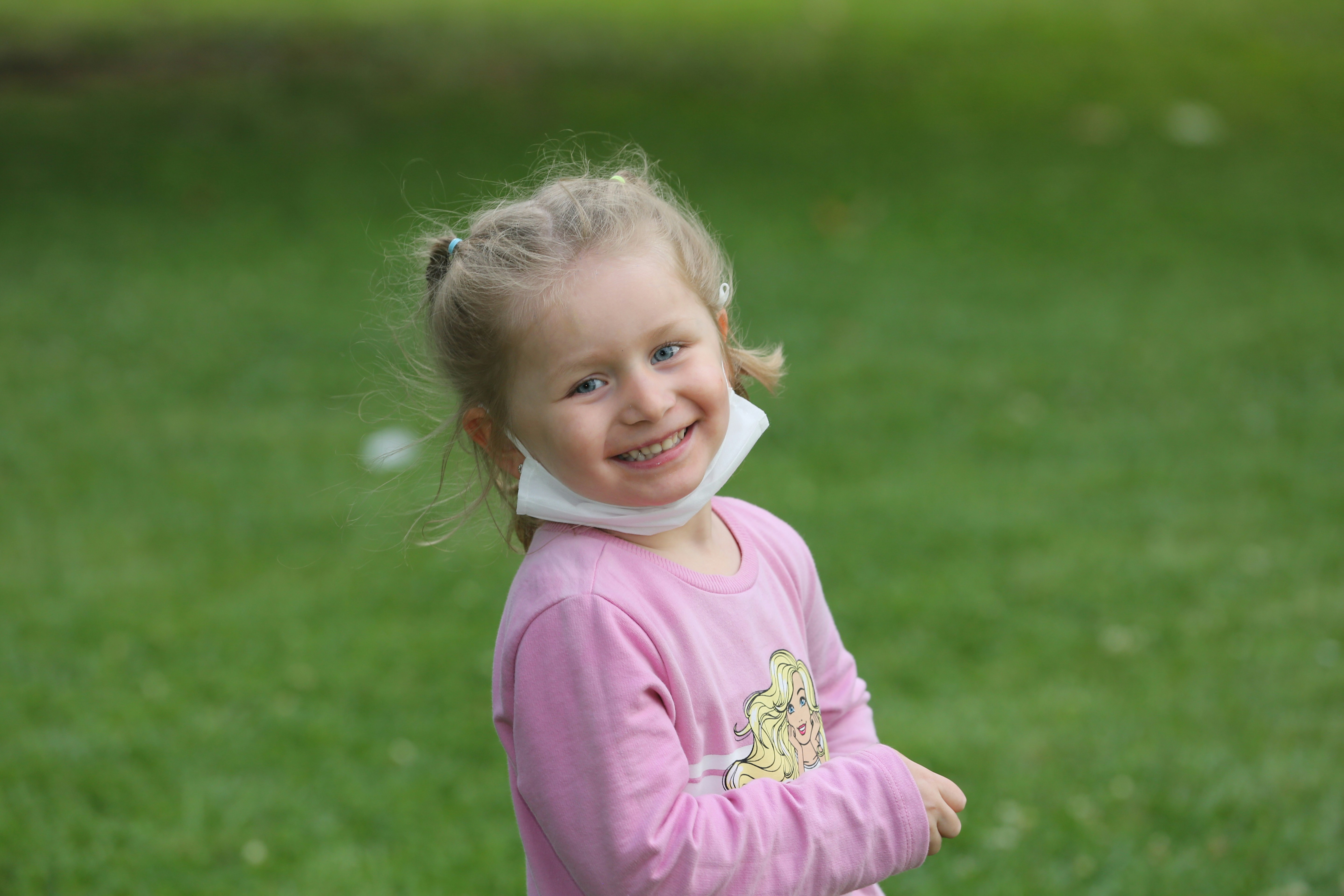 Young girl with a beaming smile wearing a mask, set against a vibrant green lawn.