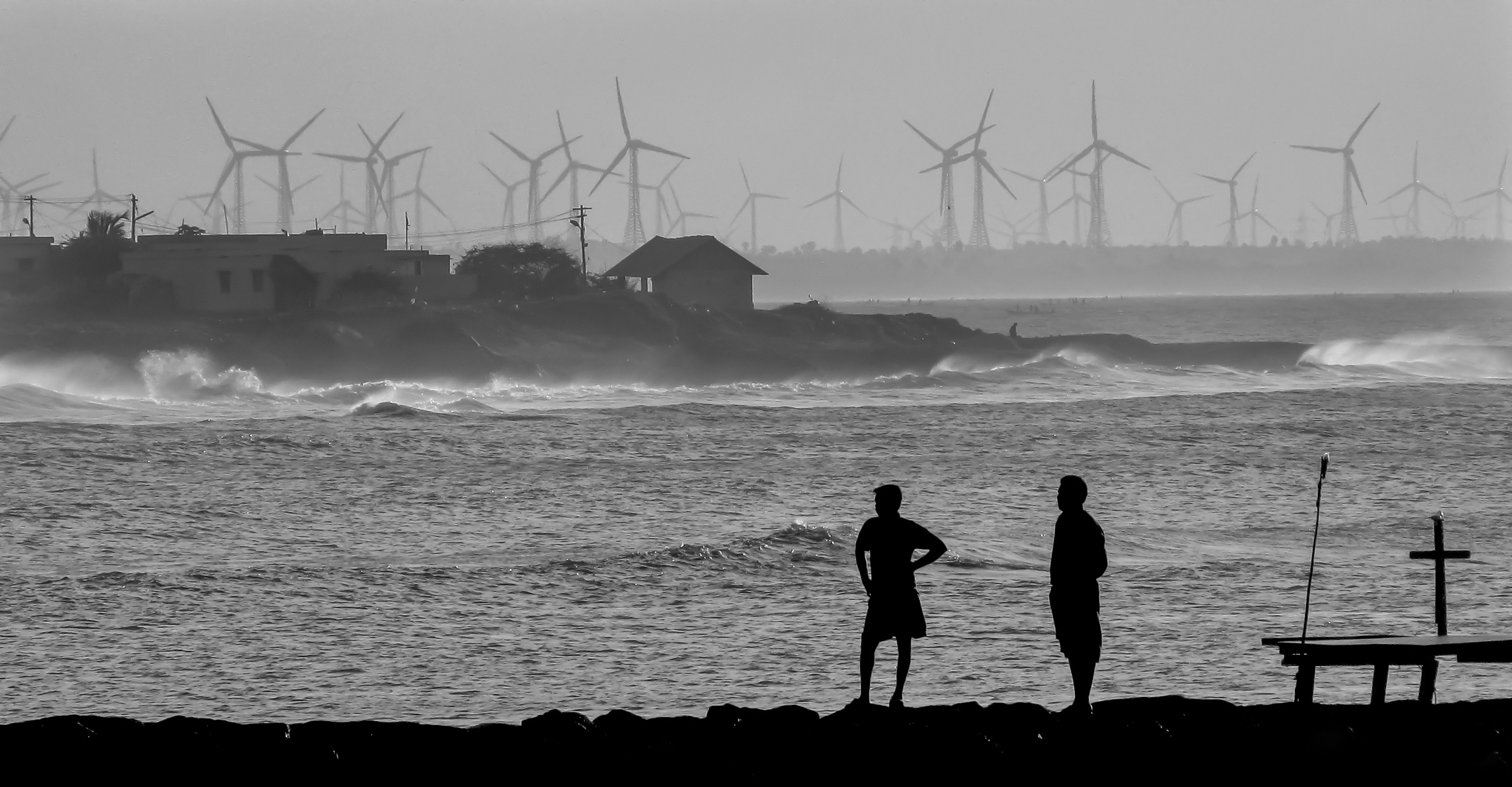 Two Silhouettes at Distant Shore as Wind Turbines Rise Behind