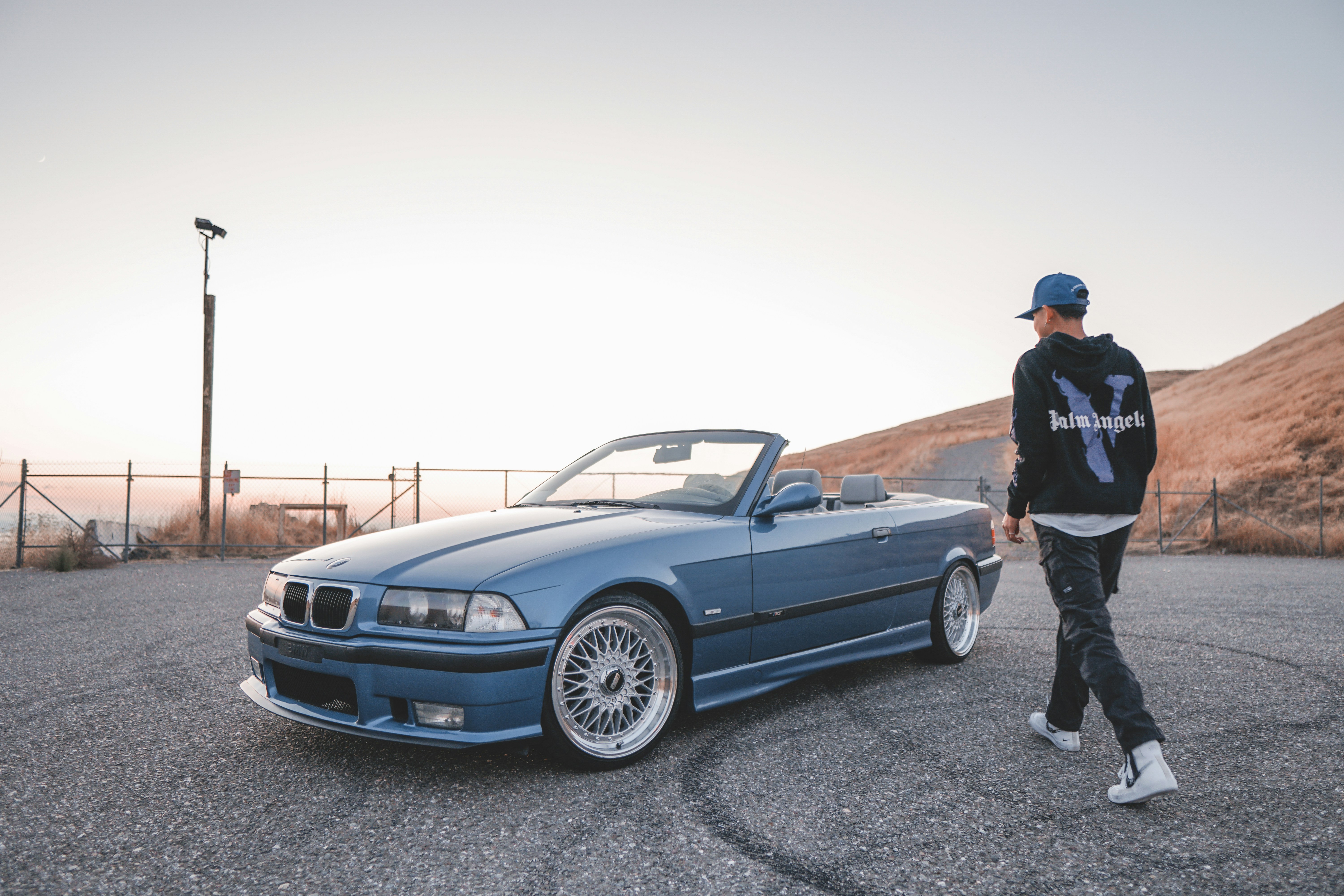 Person walking towards a blue convertible parked on a mountain road at dusk.