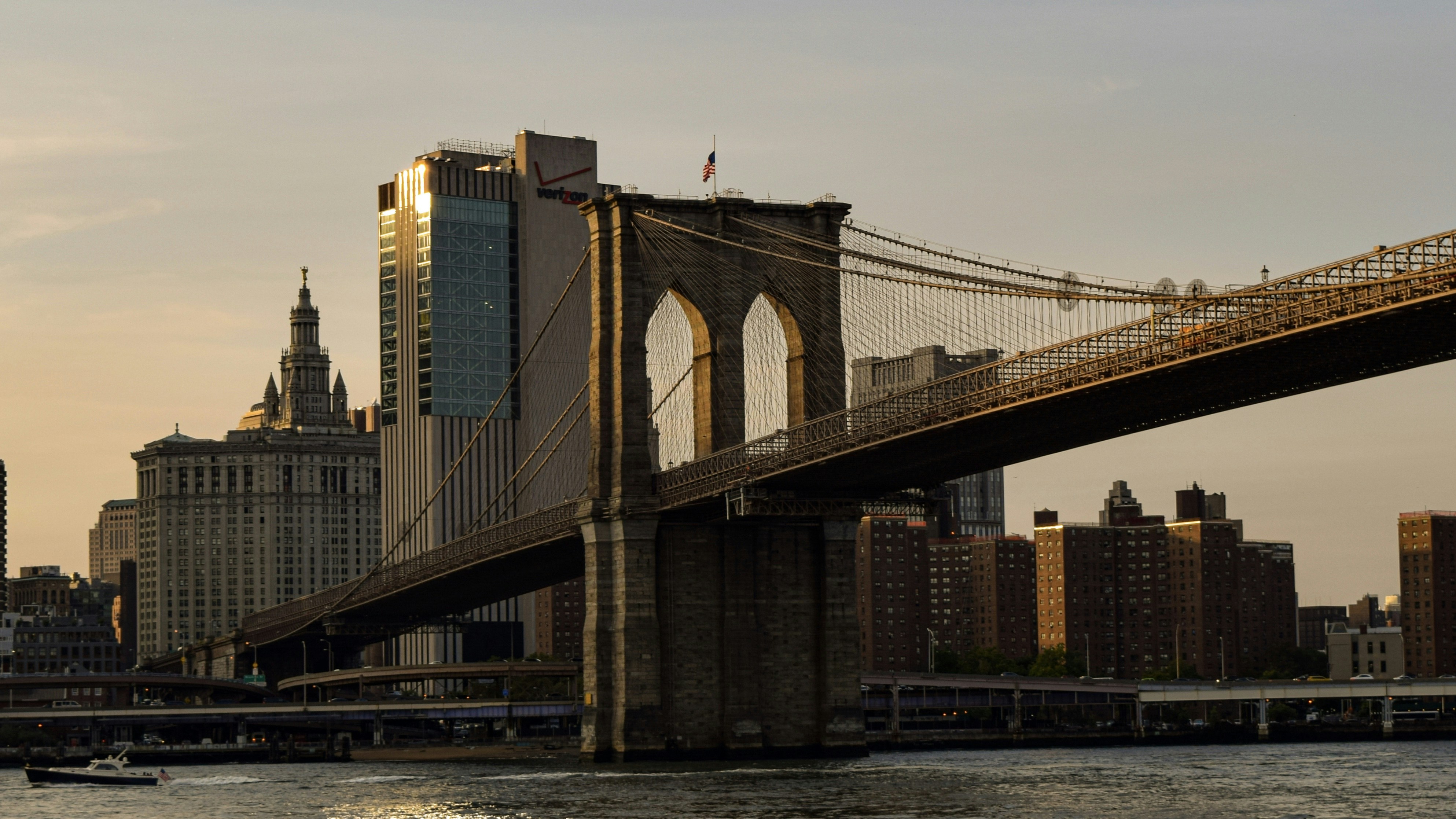 Brooklyn Bridge spans the East River against a golden sunset, with New York City's skyline in the background.