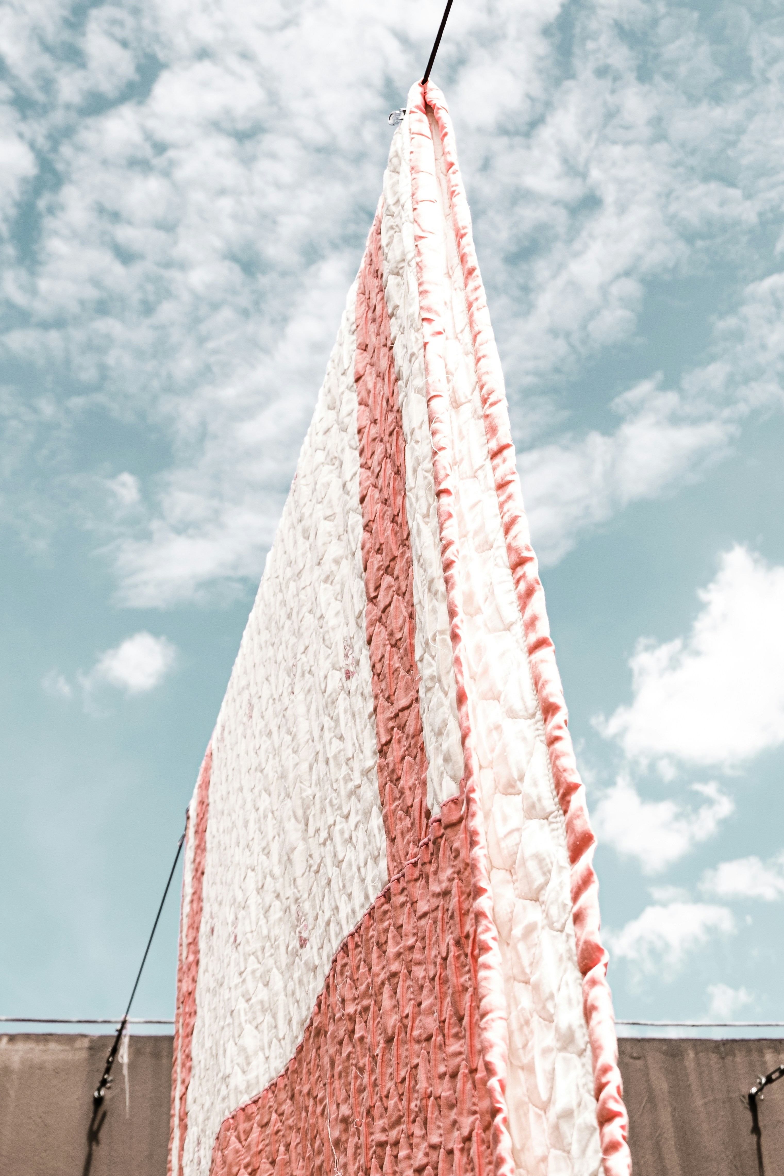 A vibrant quilt hangs against a backdrop of fluffy clouds and blue sky, showcasing intricate patterns and textures. The fabric's colors contrast beautifully with the serene atmosphere.