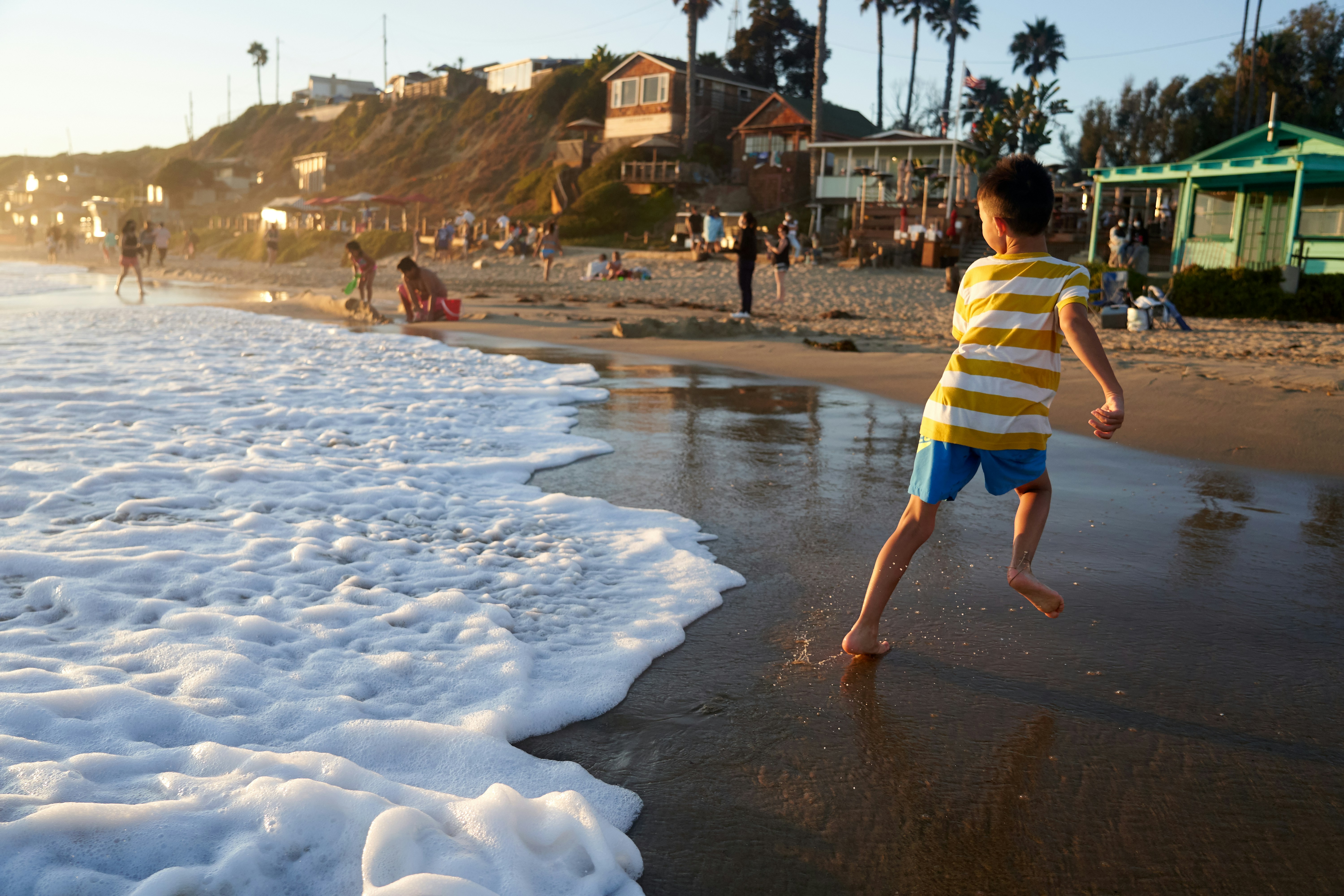 Child in striped shirt runs along the shoreline as gentle waves wash over the sand at sunset.