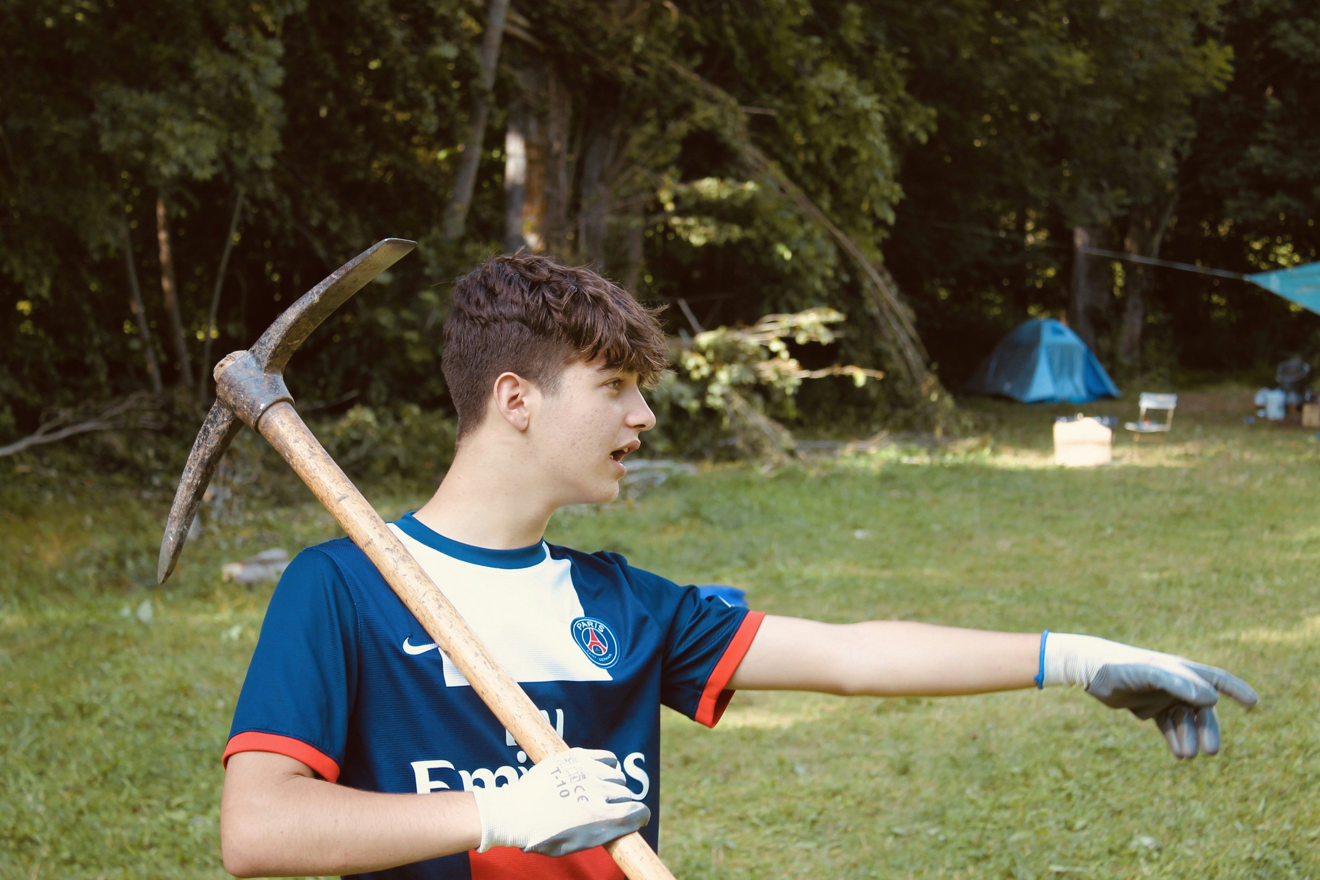 A young man in a blue sports jersey extends his arm, gripping a pickaxe at a grassy campsite, with tents and trees in the background.