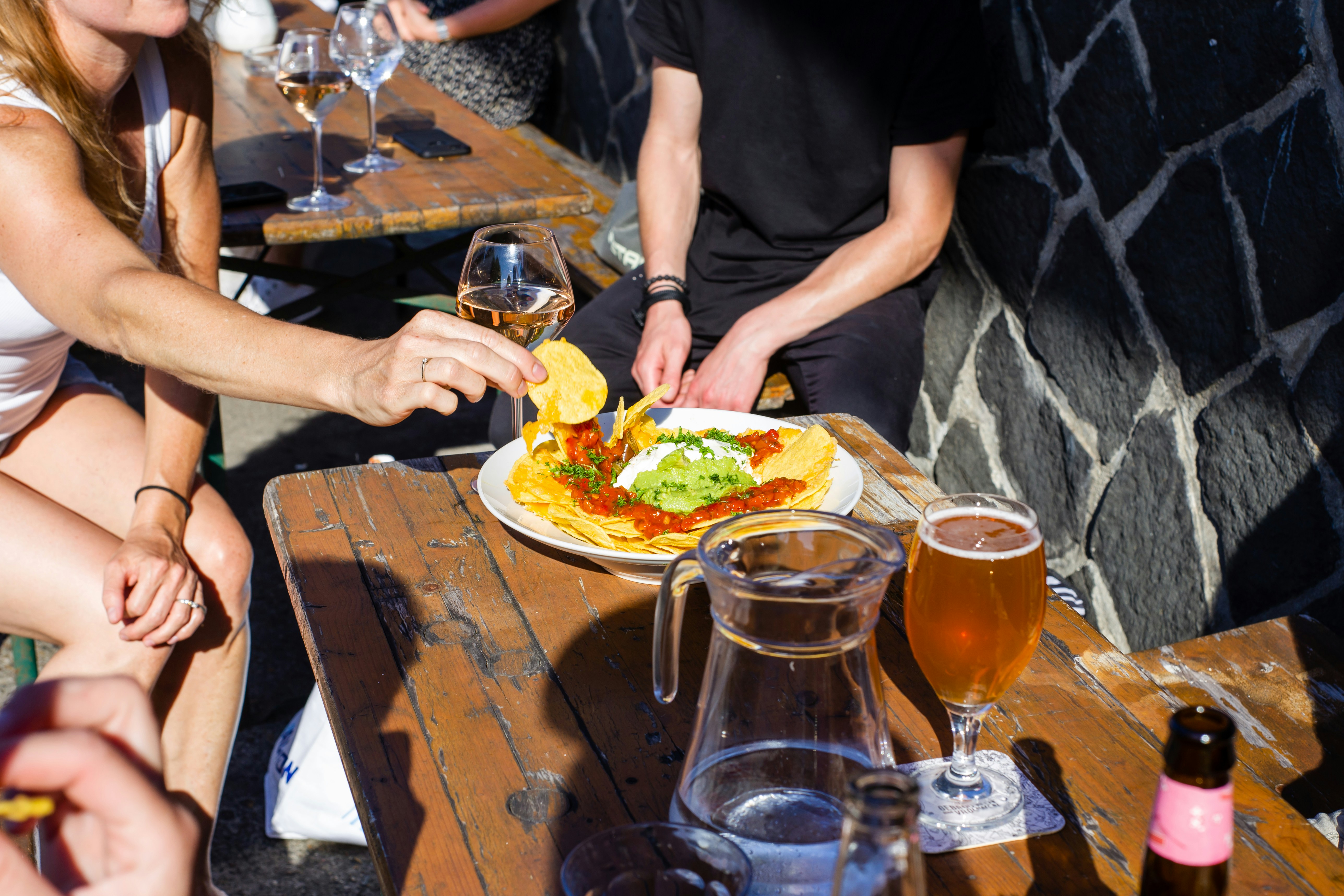 Outdoor gathering with friends sharing pizza and drinks on a wooden table under bright sunlight.