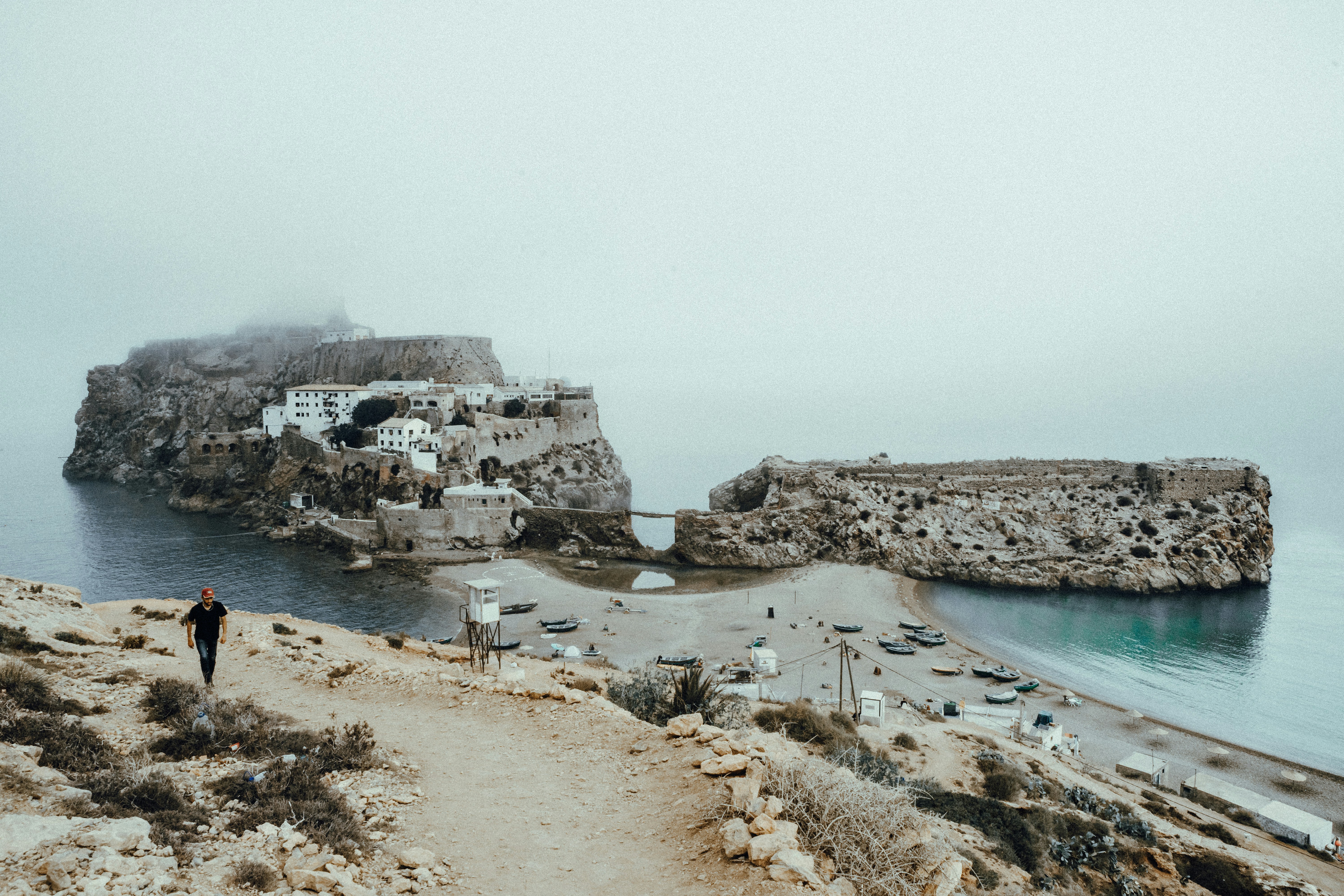 A rocky island village shrouded in fog, with boats anchored in the serene waters below. The path leading to the coast invites exploration.