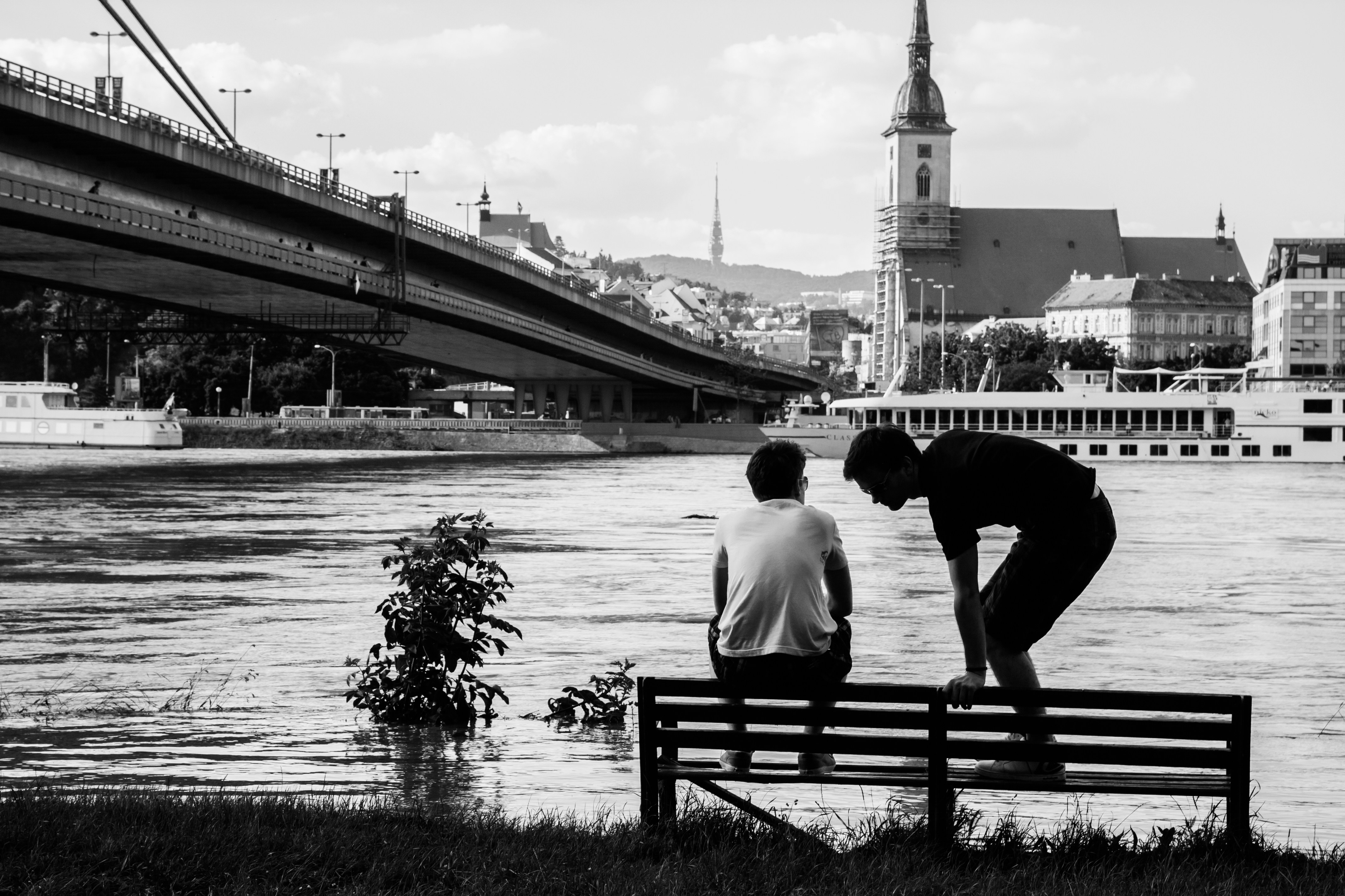 Two figures share a quiet moment on a bench by the river, framed by a bridge and distant architecture. The monochrome setting enhances the intimacy of their conversation.