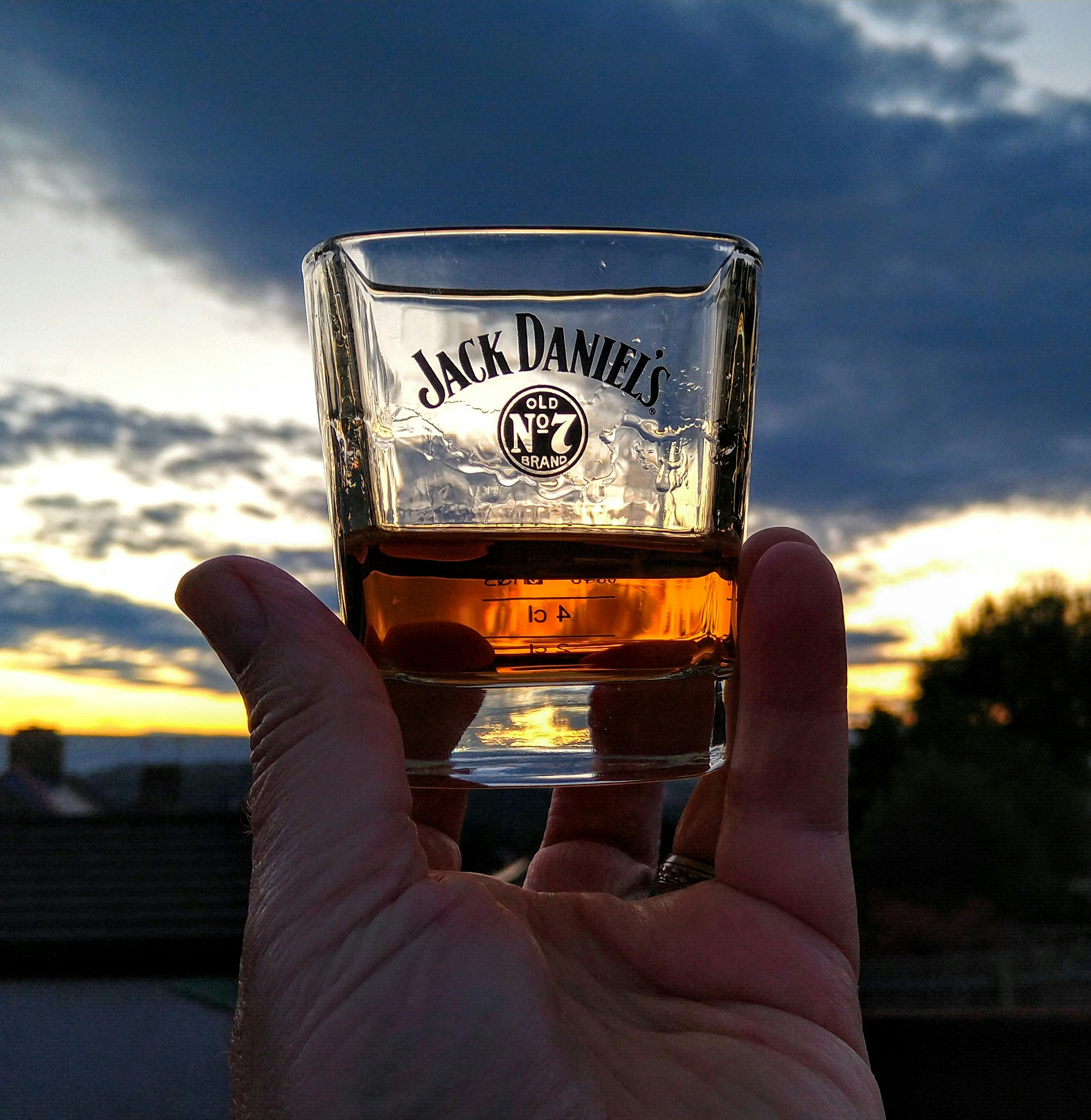 Hand holds a Jack Daniel's glass up to a sunset, amber liquid catching the light. The scene captures a warm glow against a cloudy sky and distant rooftops.