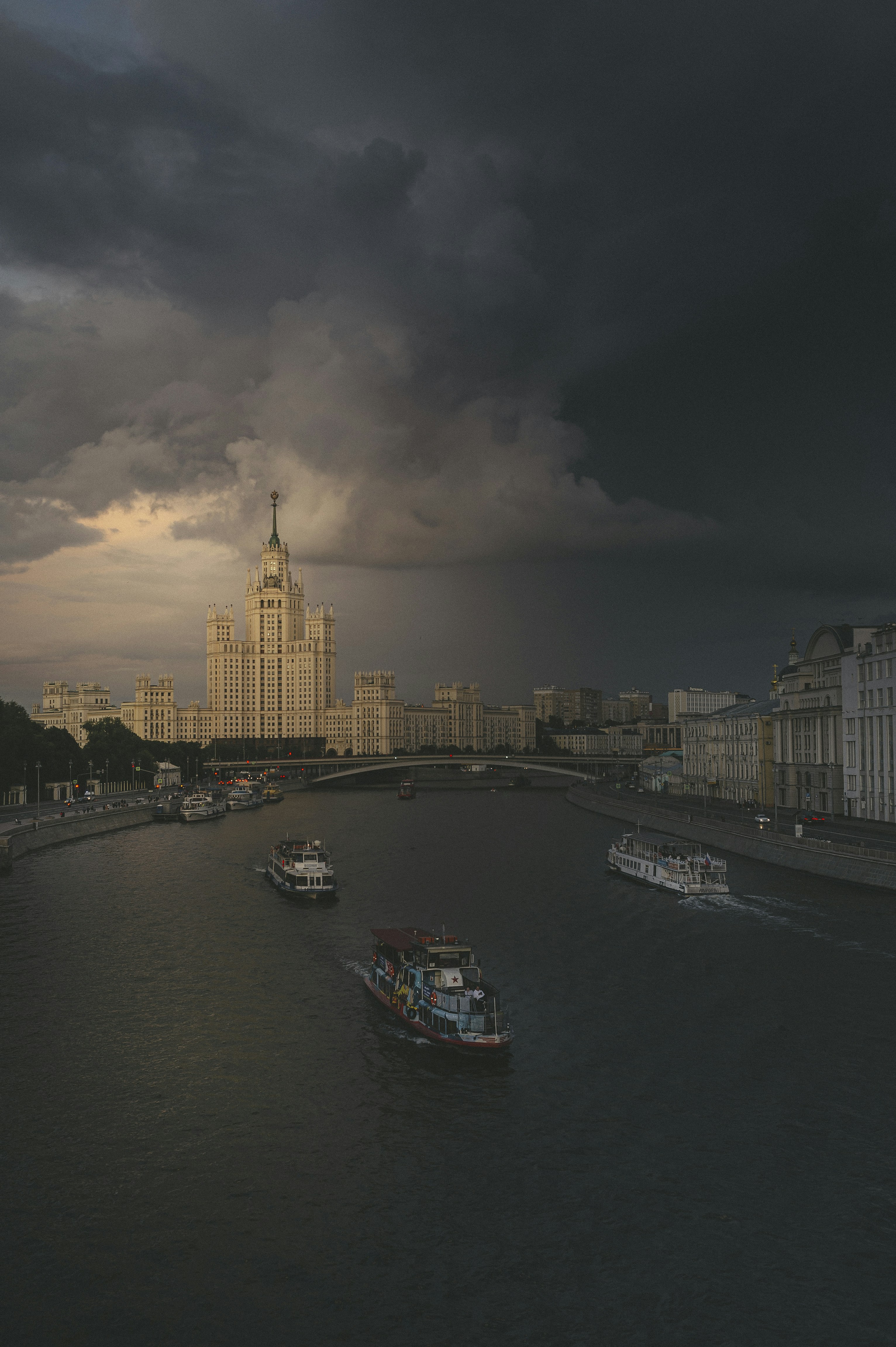 City riverfront at dusk under approaching storm clouds, featuring a tall illuminated spire rising above the skyline and boats moving along the water.
