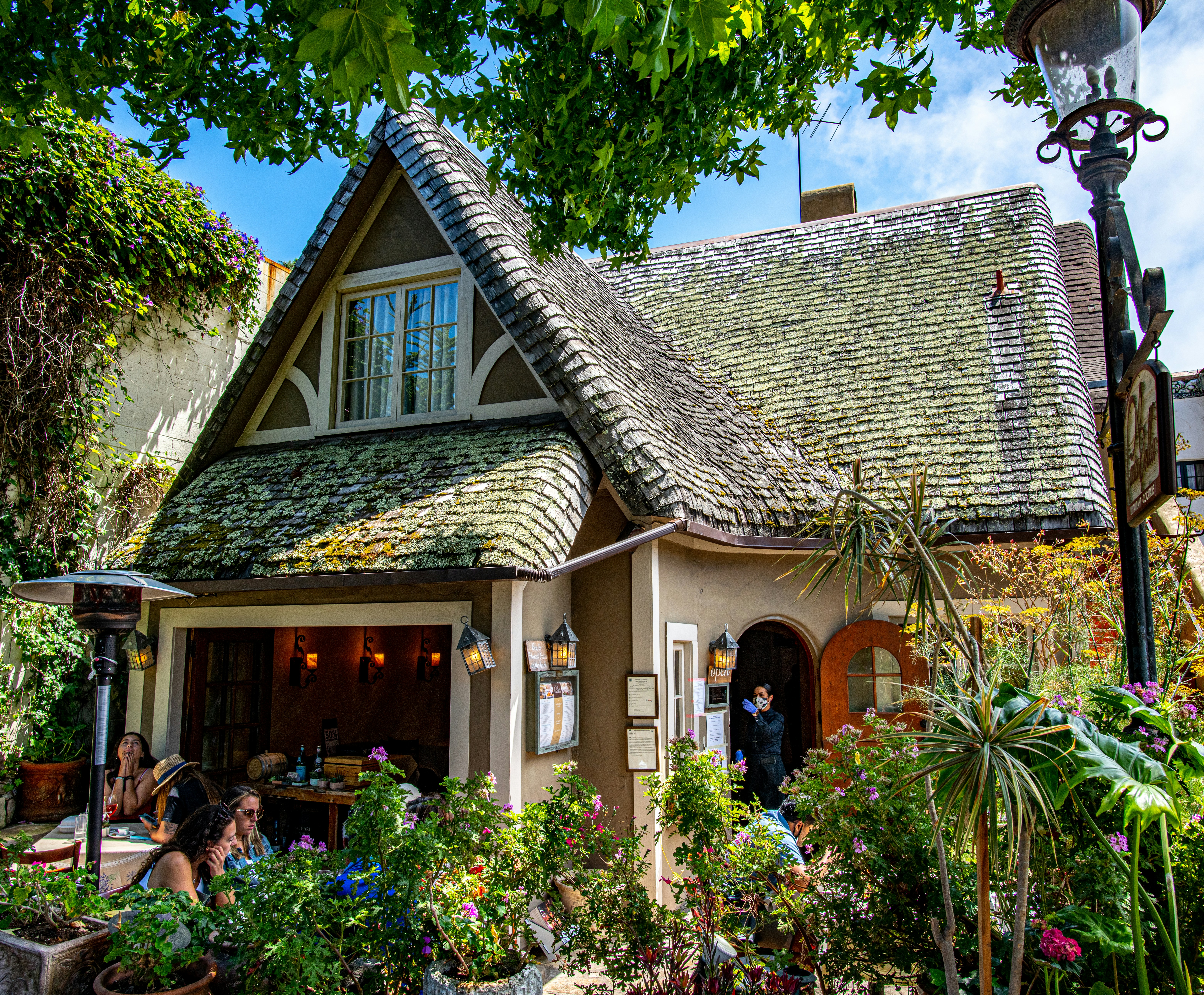 Charming cottage with a moss-covered roof surrounded by vibrant greenery and patrons enjoying the outdoor seating. The inviting atmosphere beckons for relaxation.