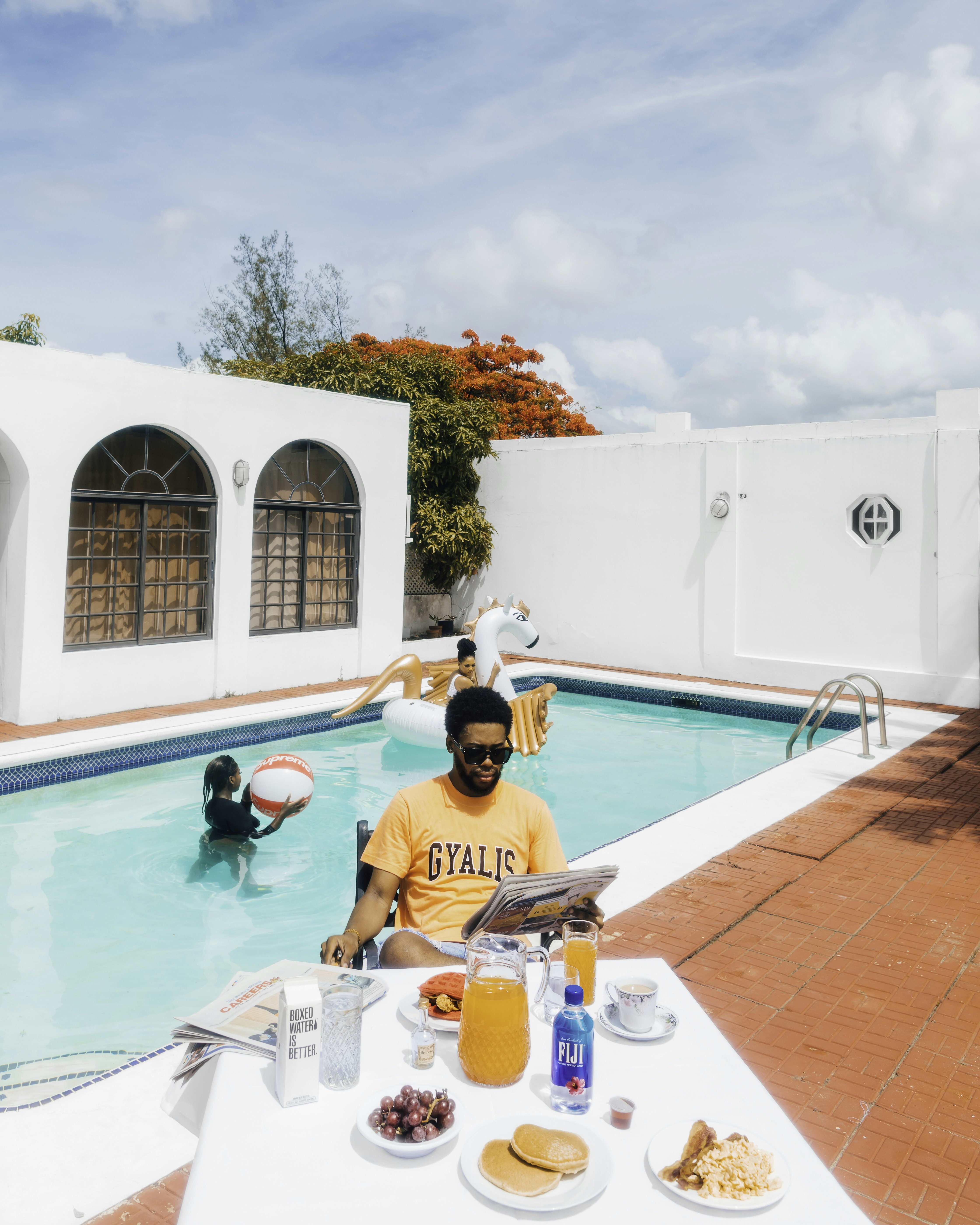 A man enjoying breakfast by the pool, surrounded by vibrant decor and playful activities. A child splashes in the background with a beach ball.