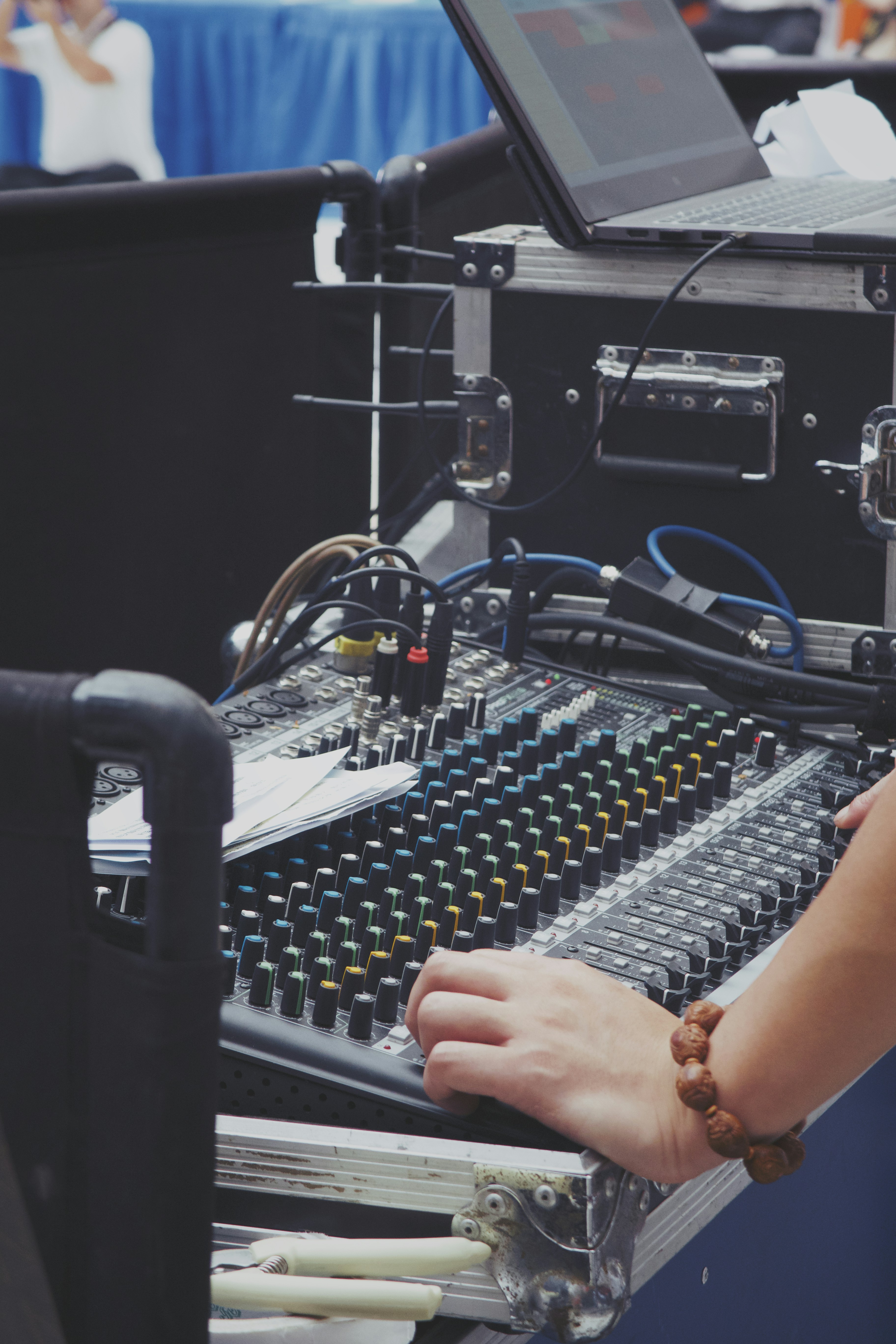 Close-up view of a technician’s hands operating a multi-channel sound mixer with a laptop on top, wires and equipment cases in the background. This photograph captures the setup used for live audio control.