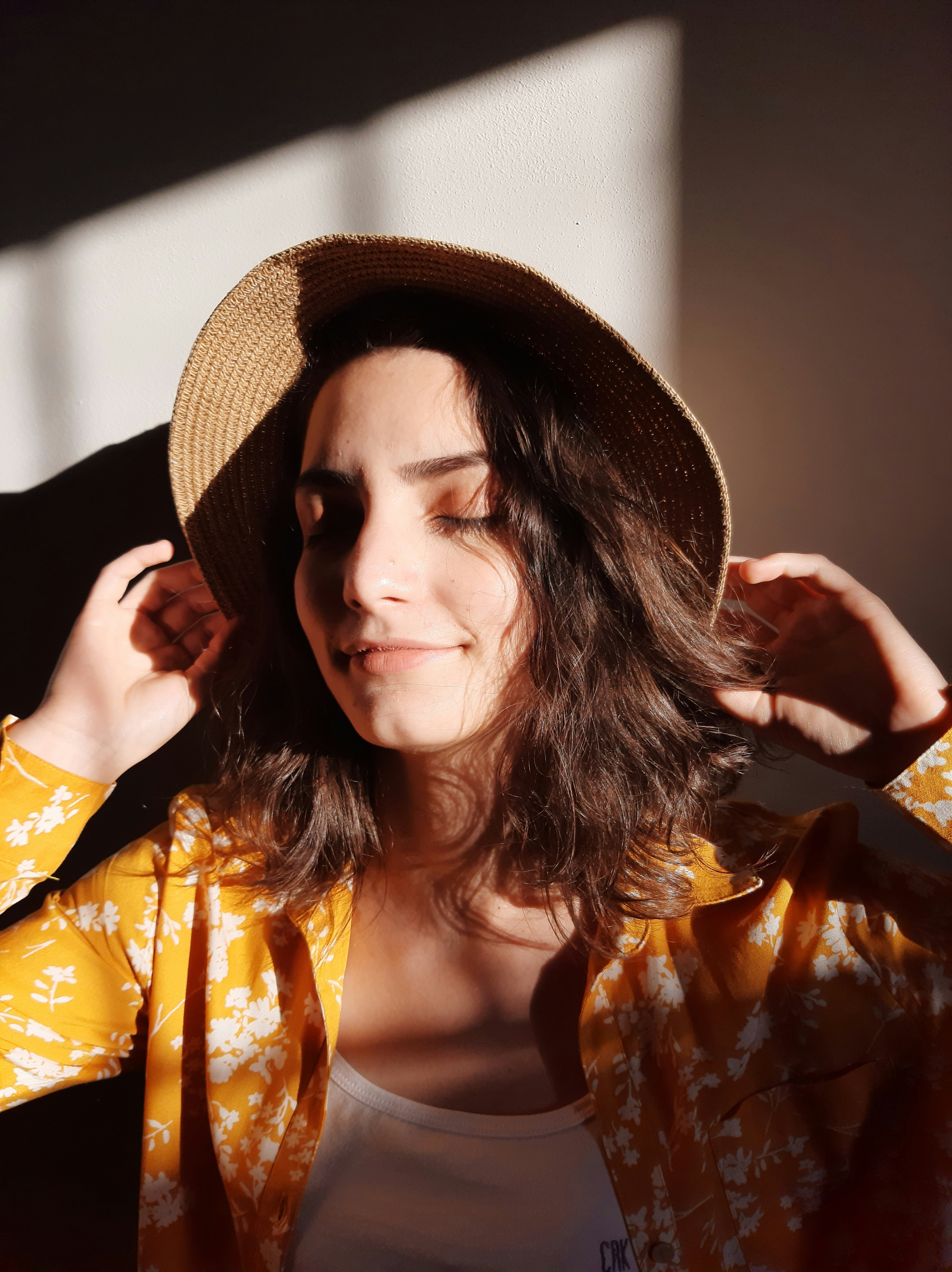 Portrait of a woman wearing a straw hat bathed in warm, directional light. Soft shadows trace her features as she adjusts her hat, with a vibrant yellow-orange shirt adding color.