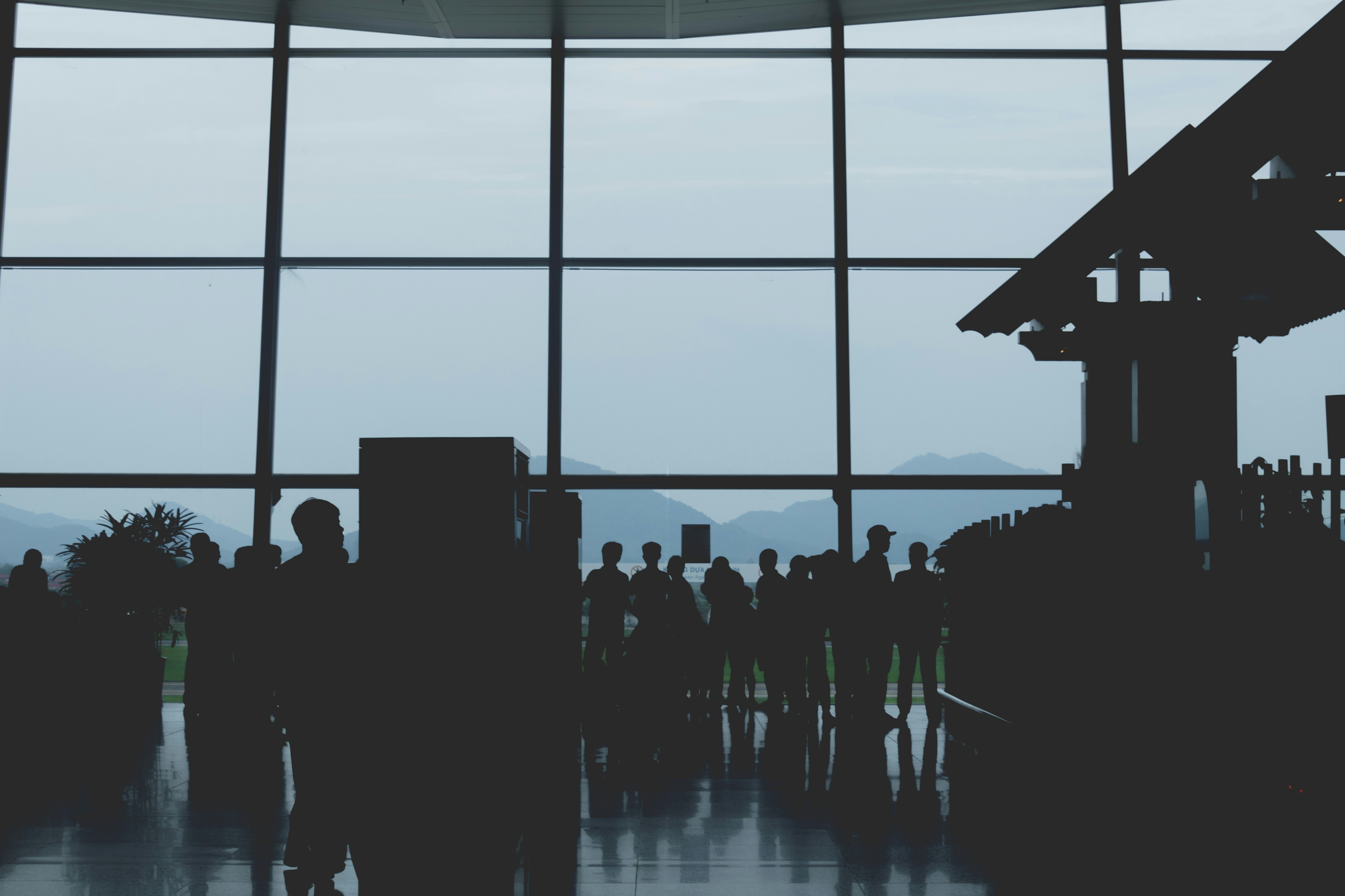 Silhouetted figures gather in front of large airport windows with an overcast sky beyond.