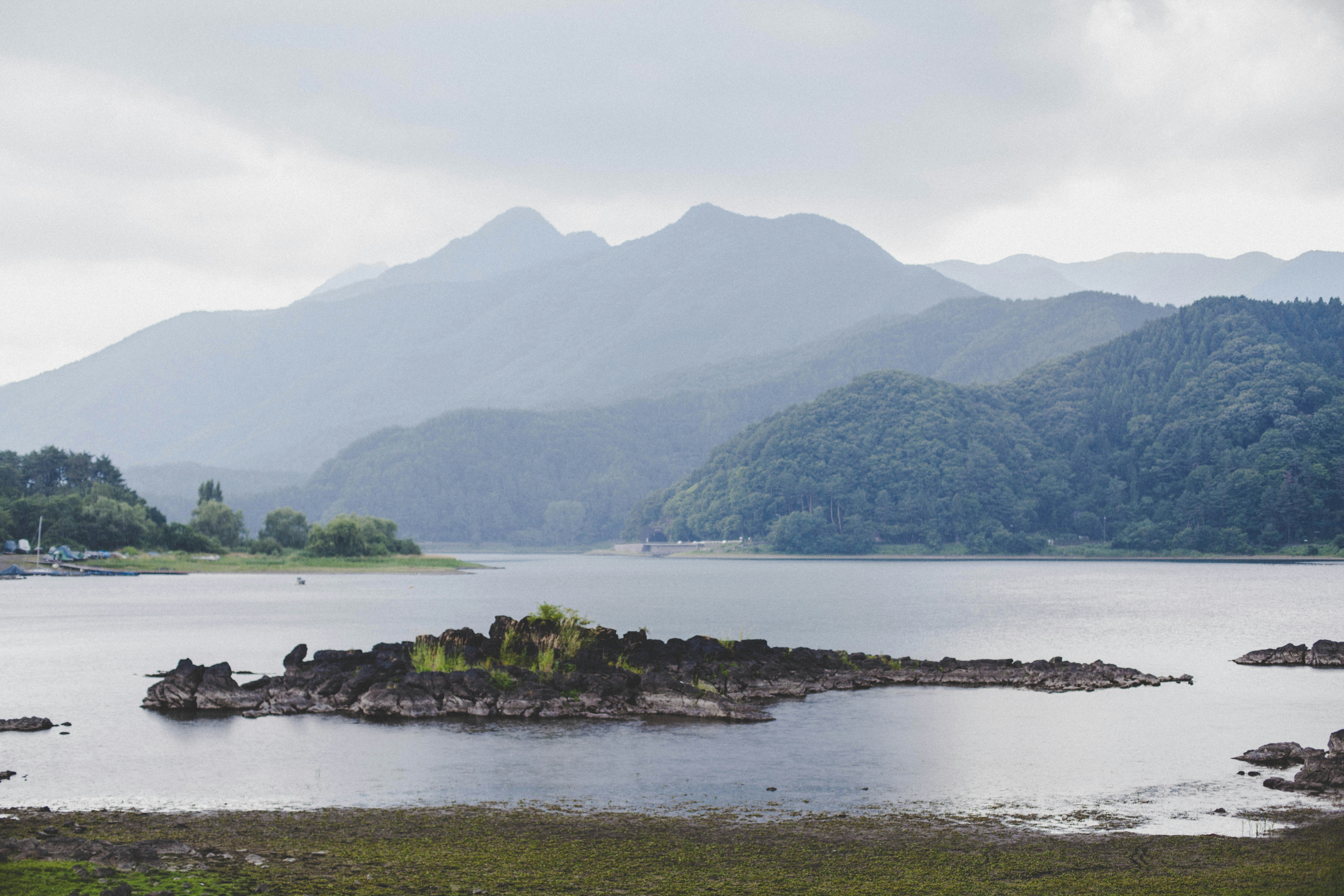 Cloud-shrouded mountains rise beyond a tranquil lake with lush greenery on the shoreline.