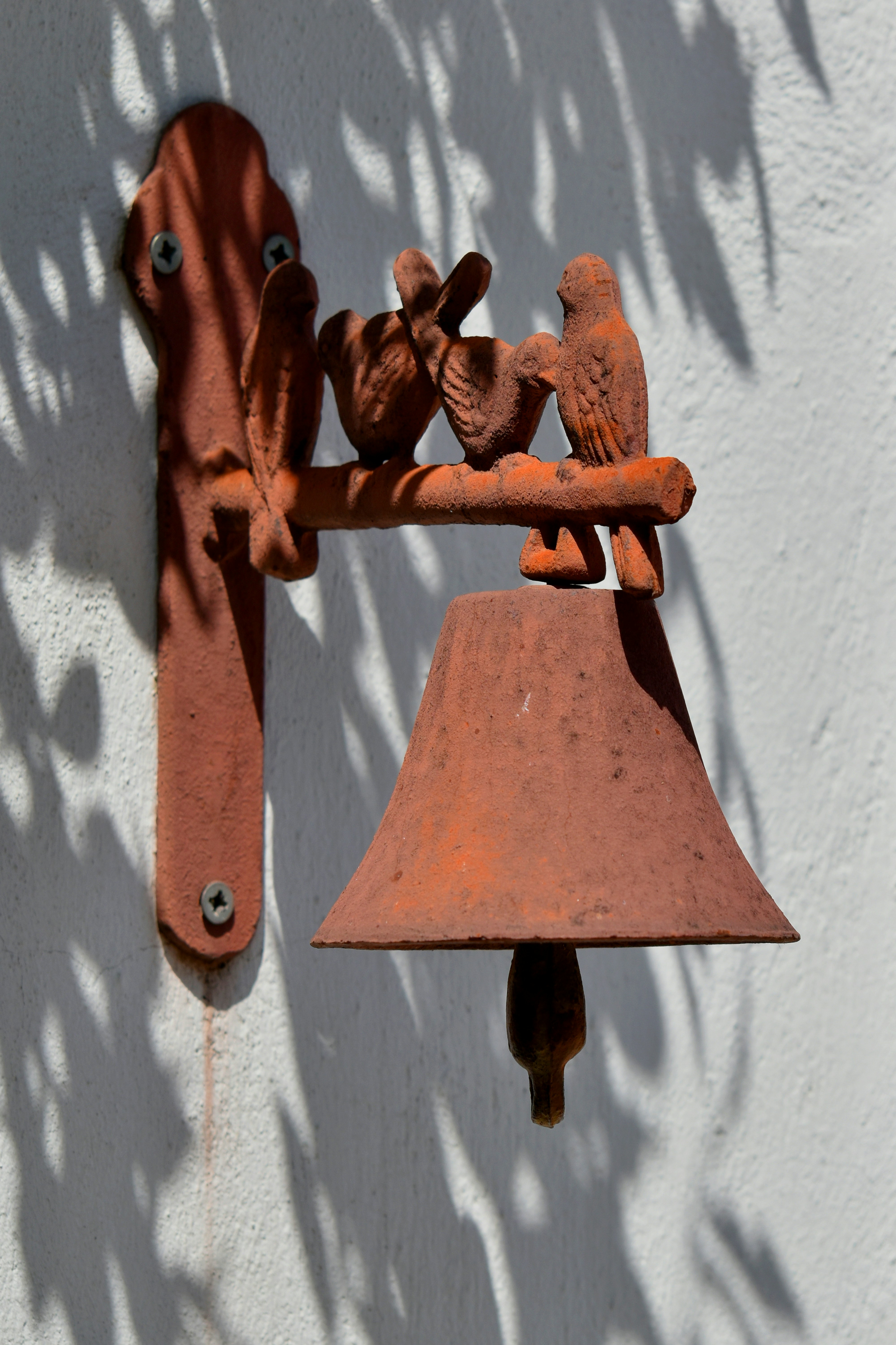 Rustic iron bell adorned with four stylized birds perched on a branch against a textured wall. Shadows play across the surface, enhancing the charm.