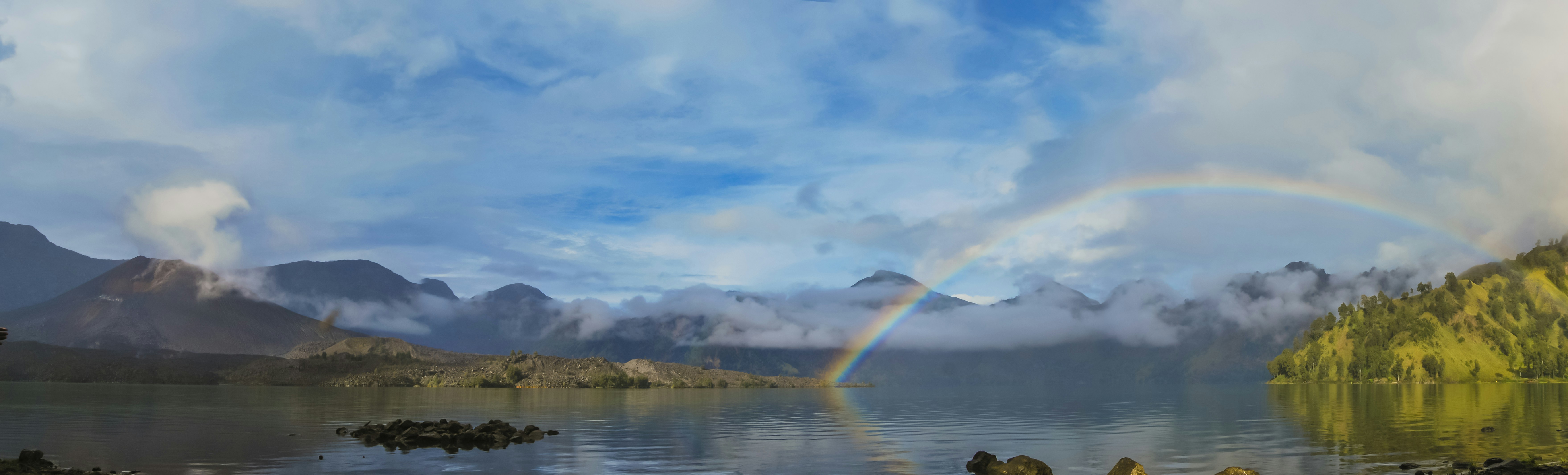 Vibrant rainbow arches over a tranquil mountain lake surrounded by misty peaks and lush greenery.