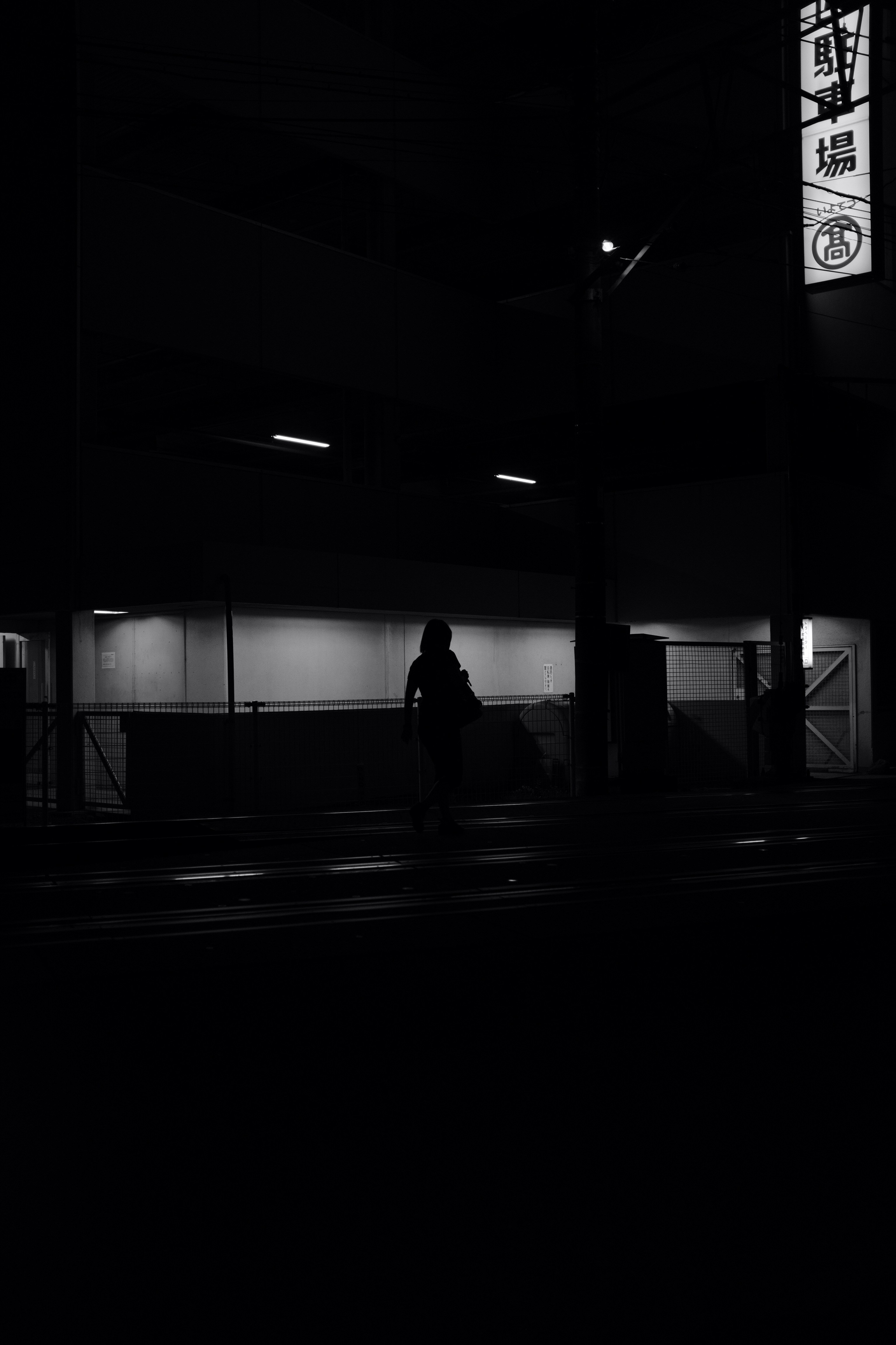 Silhouette of a figure walking alone in a dimly lit train station, with illuminated signs casting a stark contrast against the darkness.