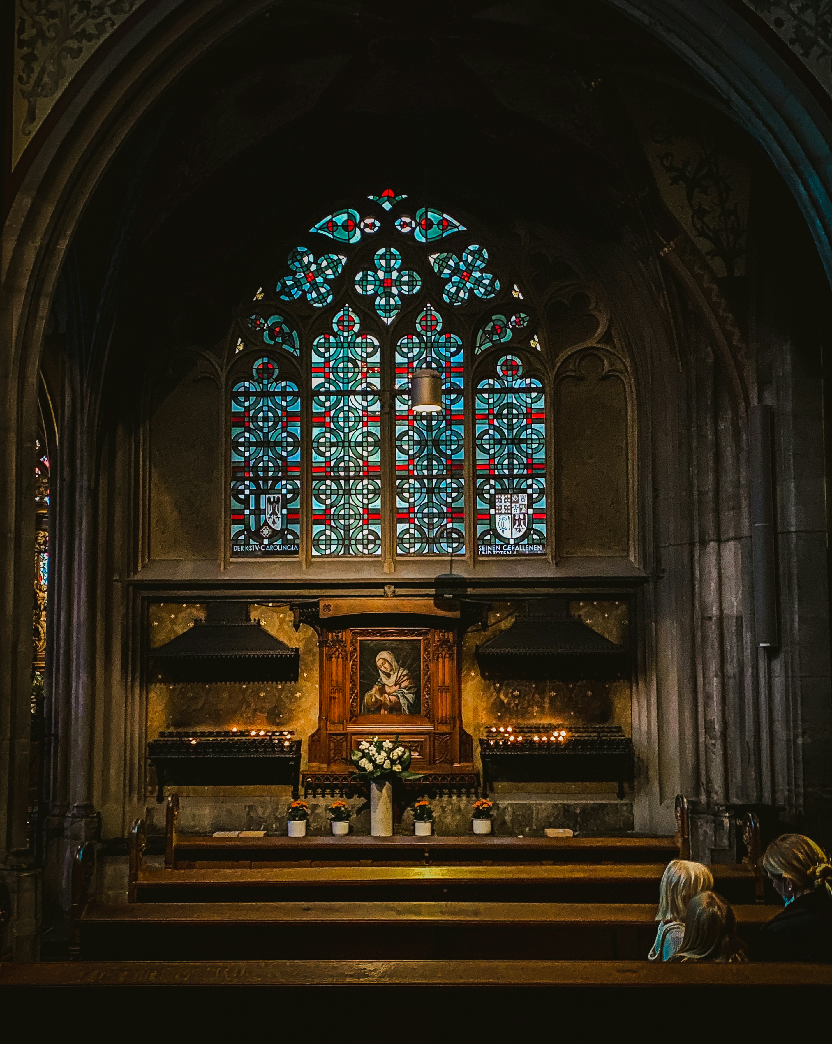 A serene chapel interior featuring a beautifully adorned altar with candles and a floral arrangement, framed by intricate stained glass windows. Soft light filters through, creating a tranquil atmosphere.