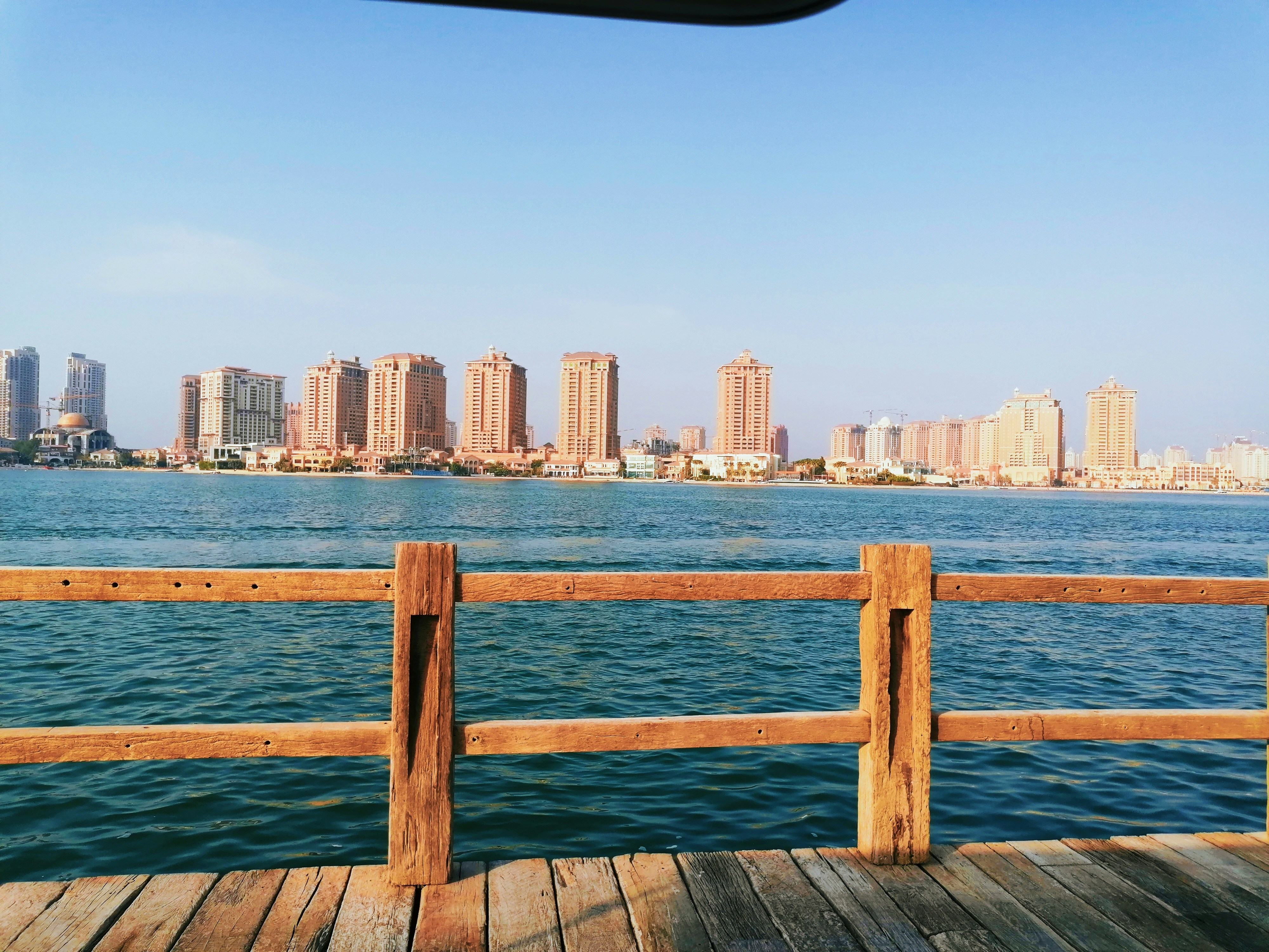 Wooden railing overlooking a calm bay with a skyline of modern high-rise buildings under a clear blue sky.