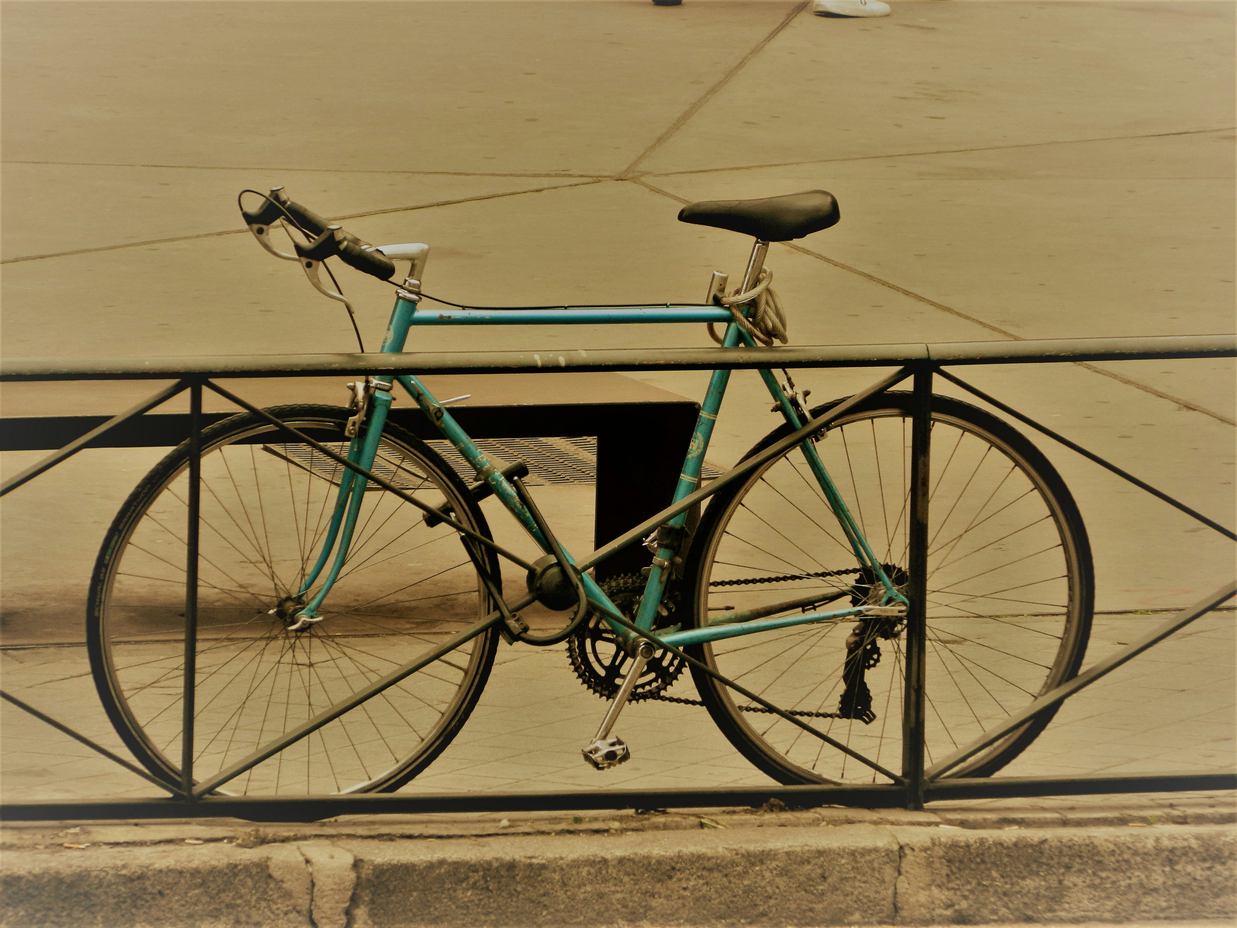 A turquoise bicycle rests against a railing on a city sidewalk, showcasing its vintage charm amidst a muted urban backdrop.