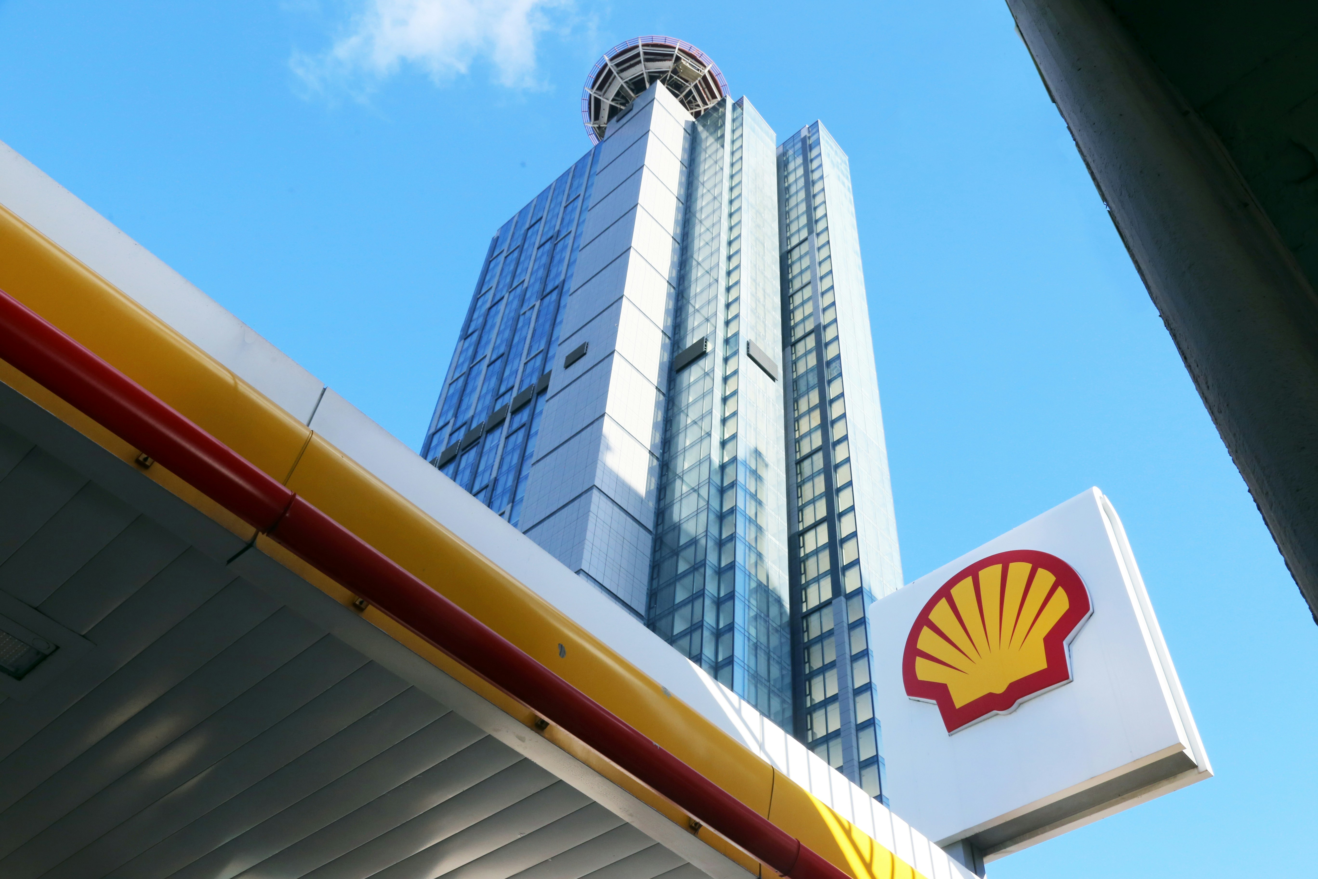 A towering skyscraper looms over a Shell gas station, showcasing contemporary architecture under a clear blue sky.