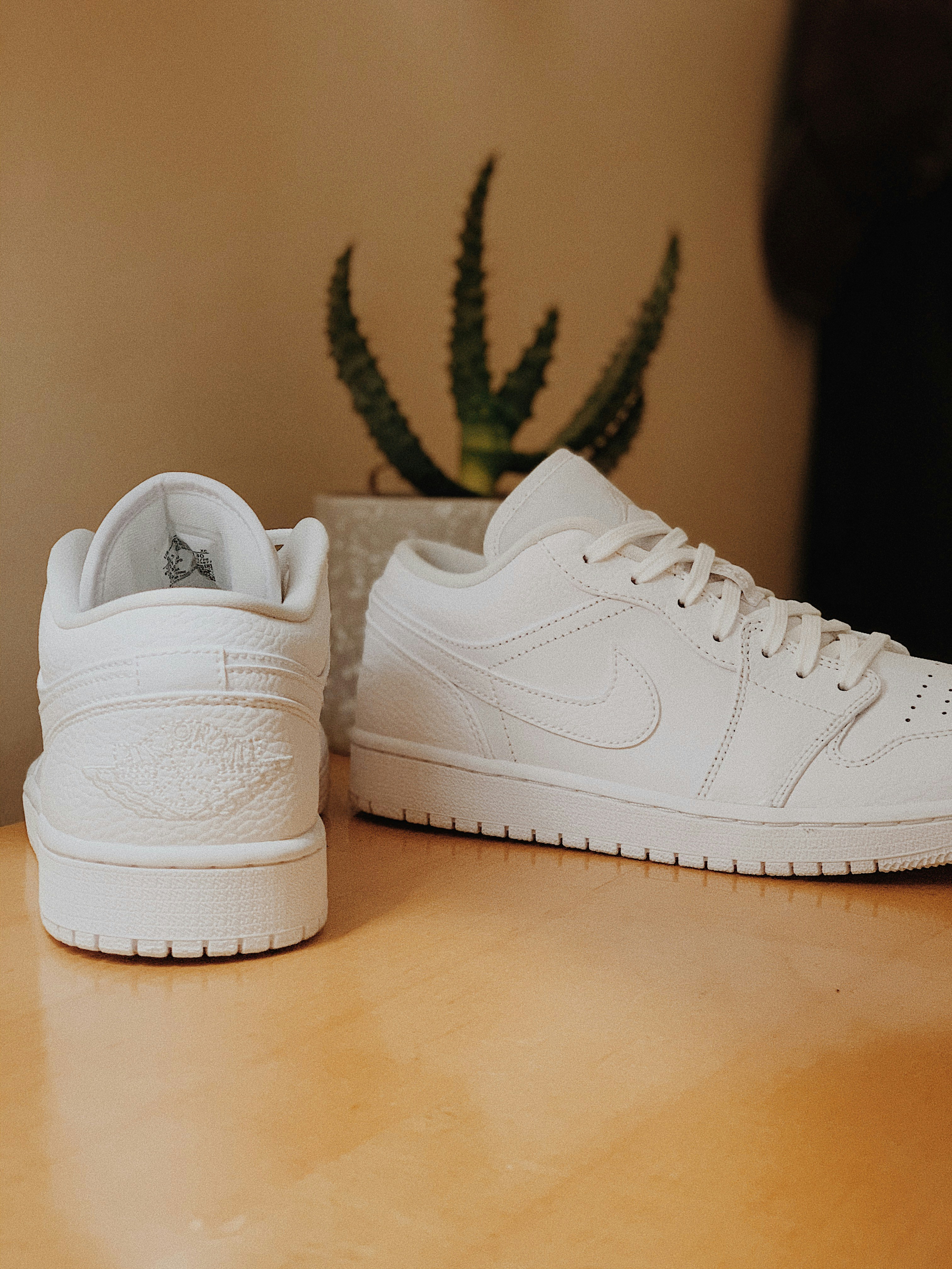 Two pairs of white sneakers displayed on a wooden surface with a potted plant in the background.