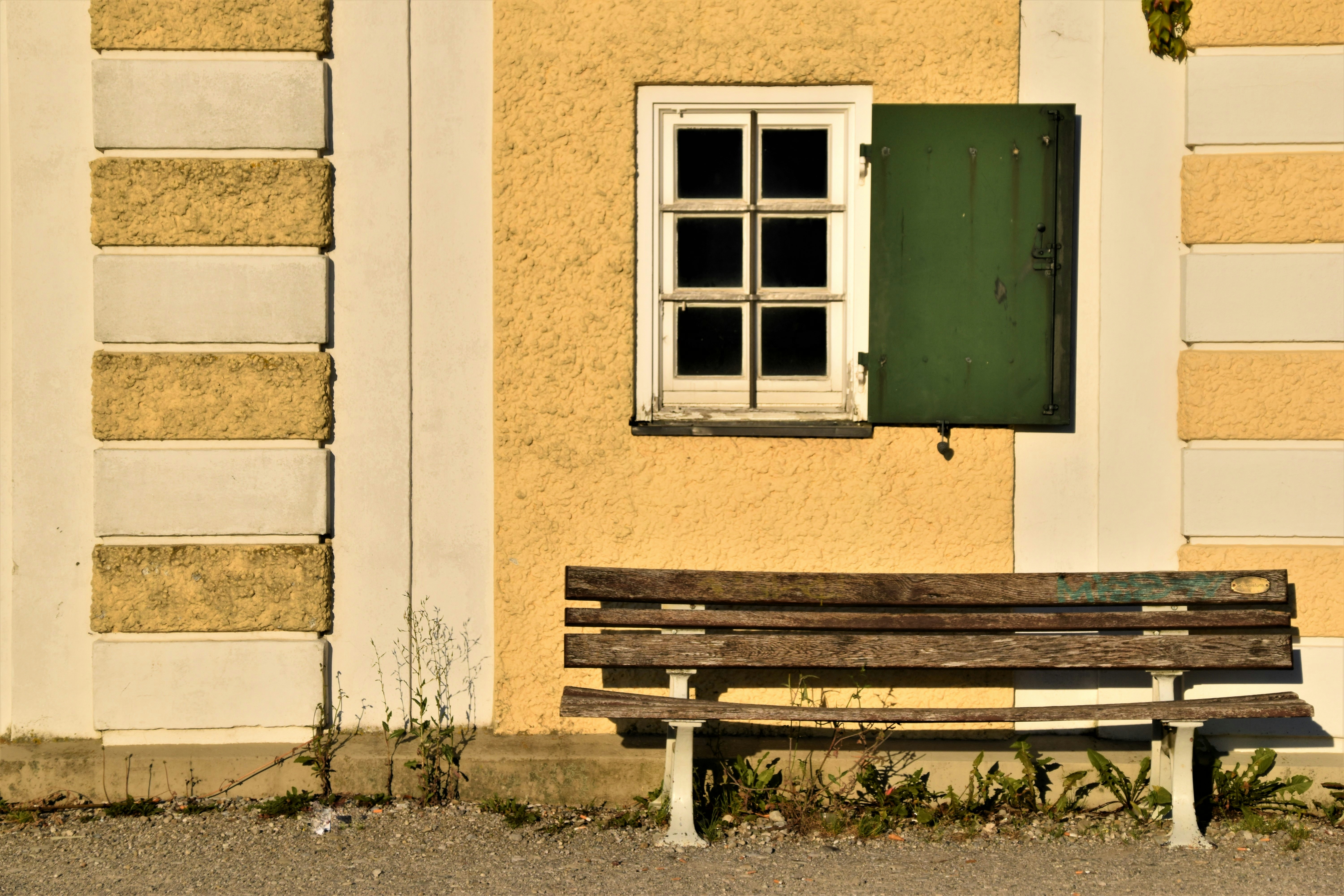 Wooden bench positioned against a textured wall with a green window shutter. The scene evokes a sense of quiet contemplation.