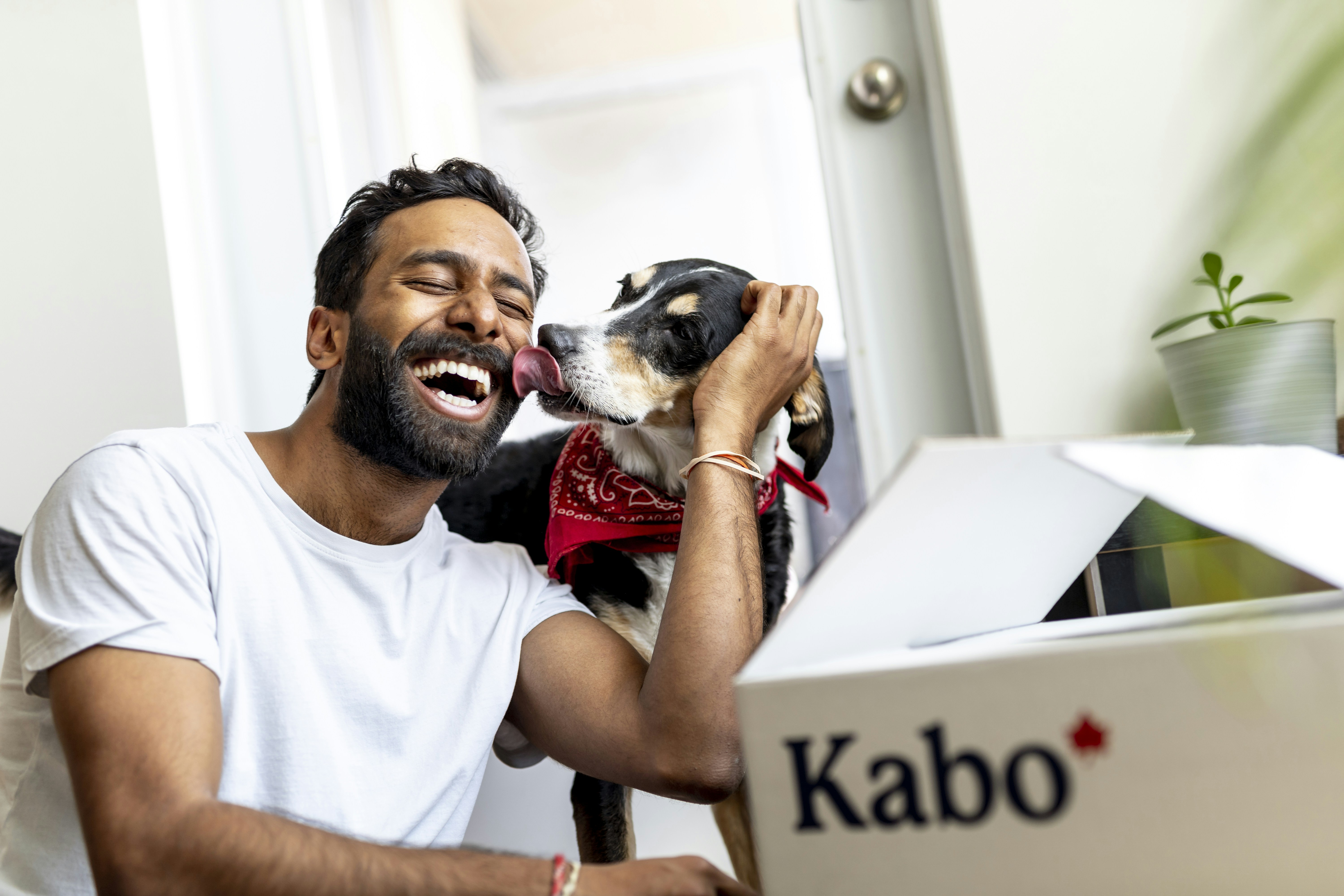 Man joyfully interacting with a dog, sharing a moment of affection and excitement over a delivery box. The dog is playfully licking the man's face.