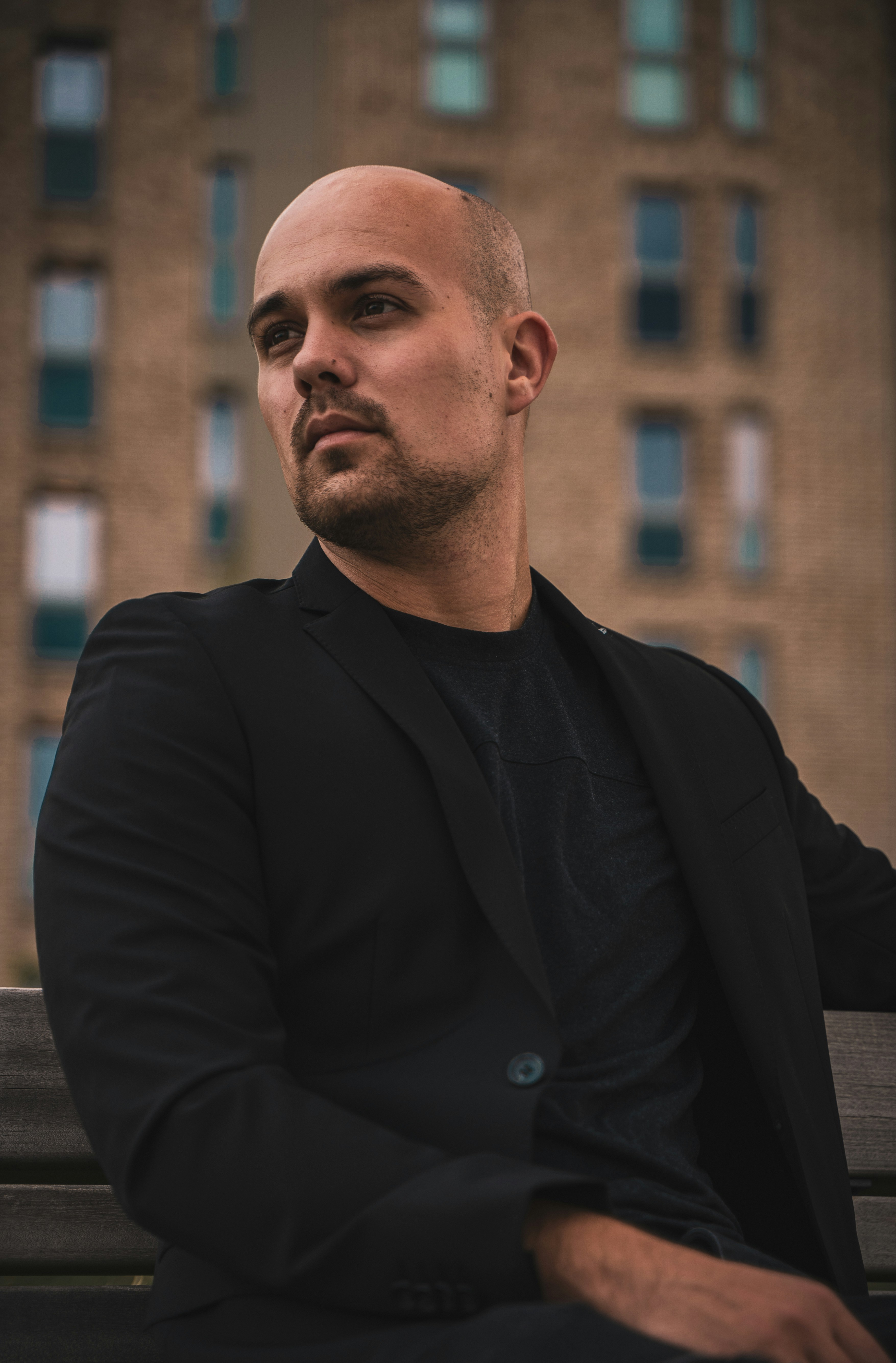 A man in a tailored black suit sits pensively on a bench, with a blurred urban backdrop emphasizing his contemplative expression.