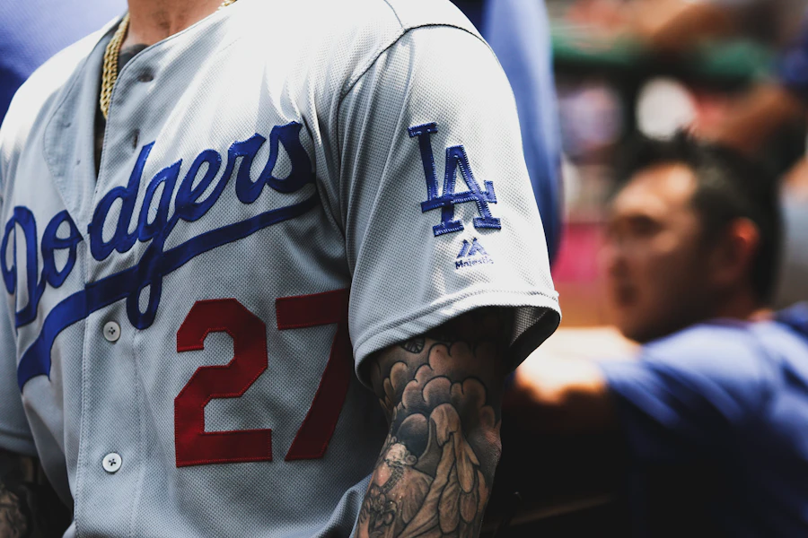 Dodgers jersey number 27 with visible LA logo patch, gold chain and detailed forearm tattoo rendered with shallow depth-of-field and strong subject isolation.