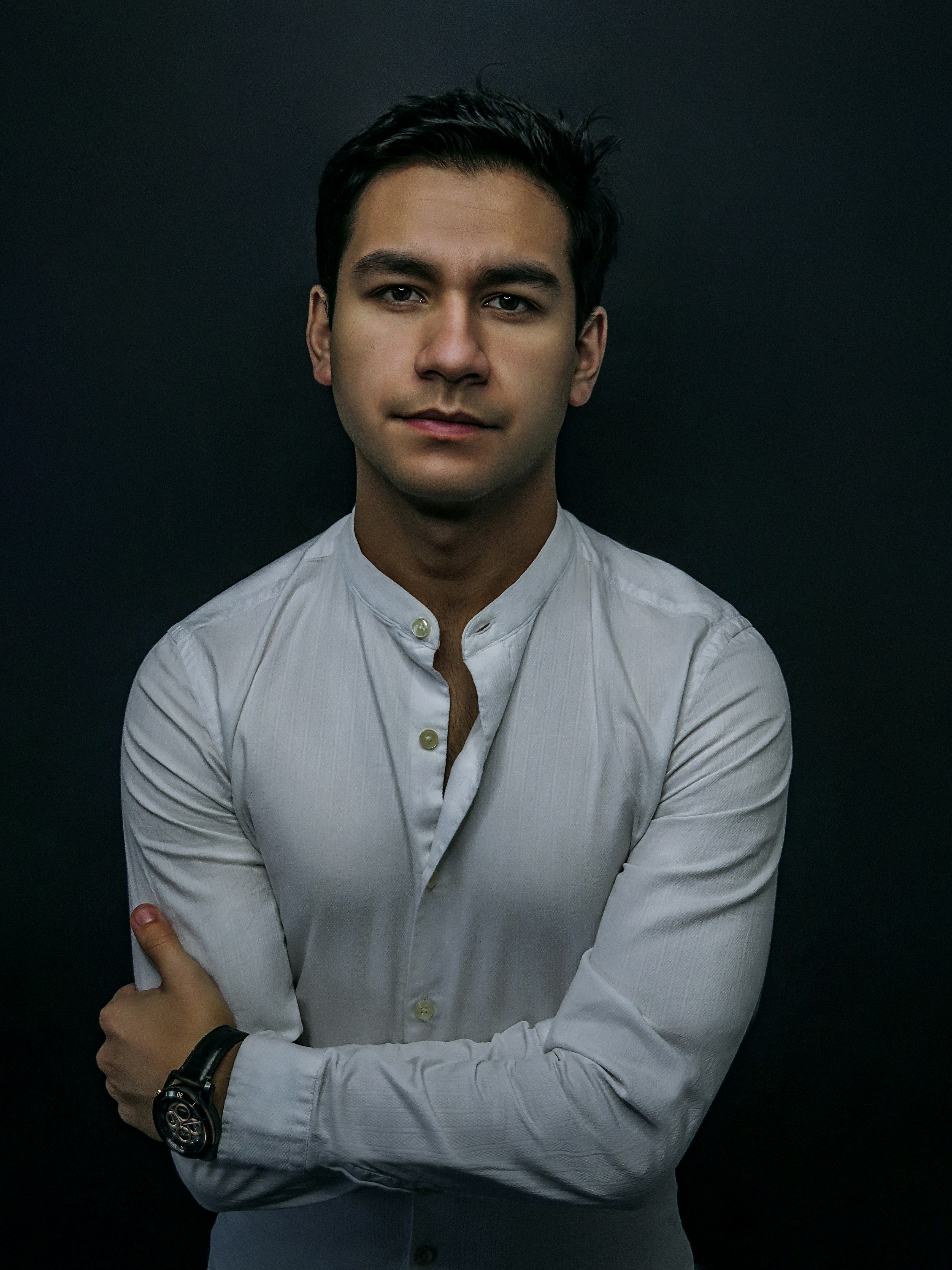 A young man in a white shirt with a watch, standing confidently against a dark background.