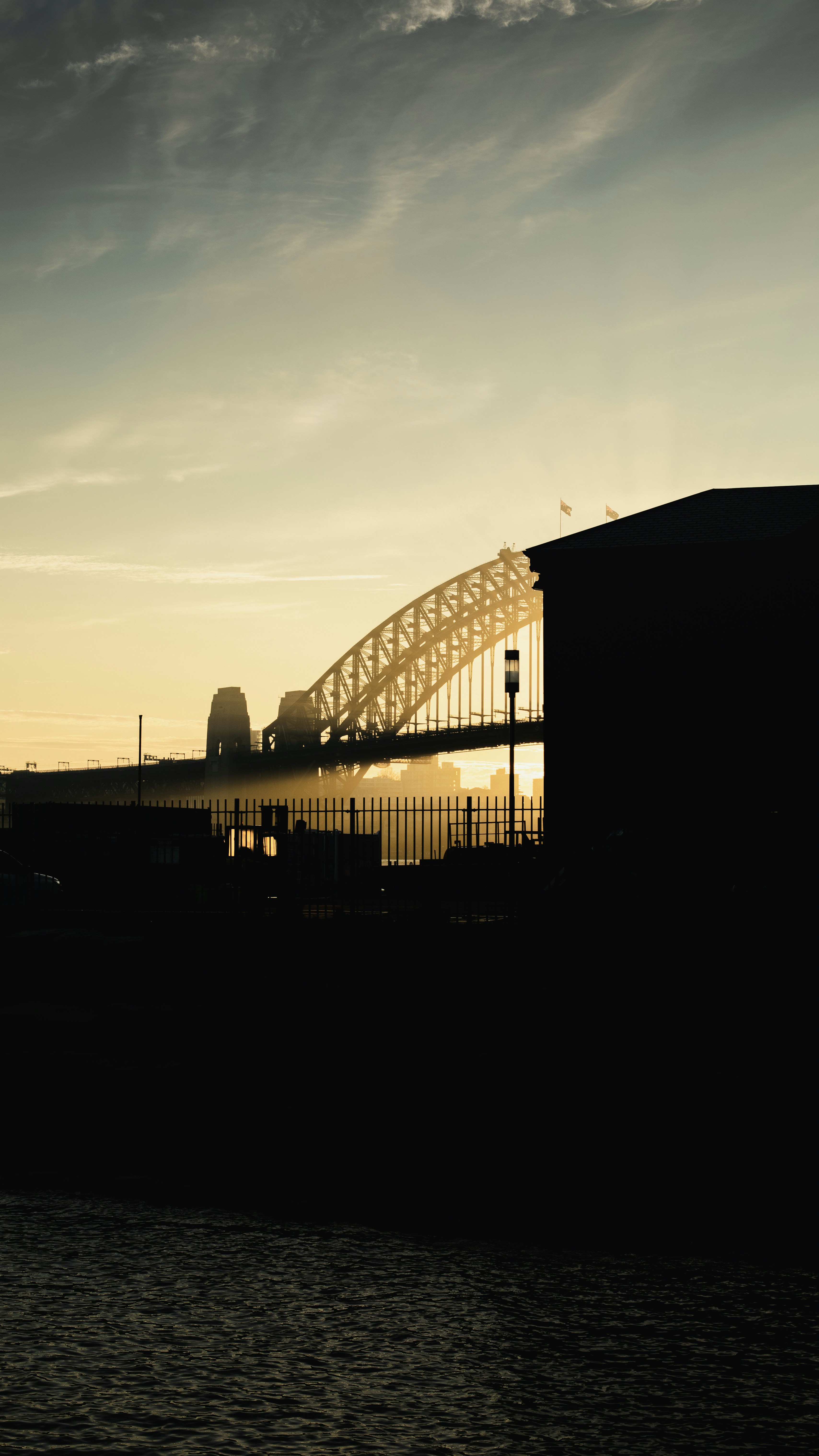 Silhouette of a bridge against a dusky sky, framed by industrial structures and gentle water reflections.