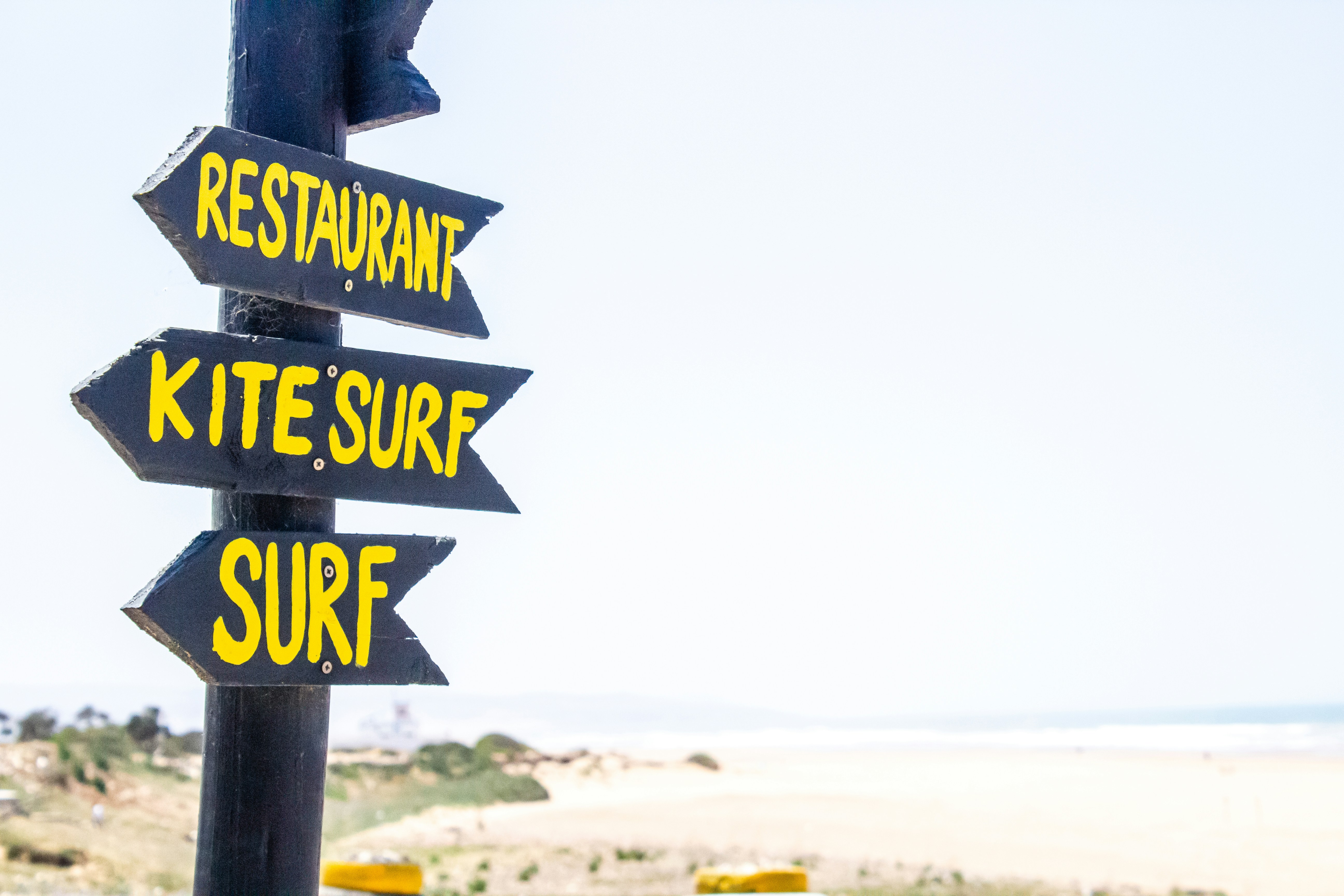 Directional signs pointing to restaurant, kite surf, and surf activities against a bright beach backdrop.